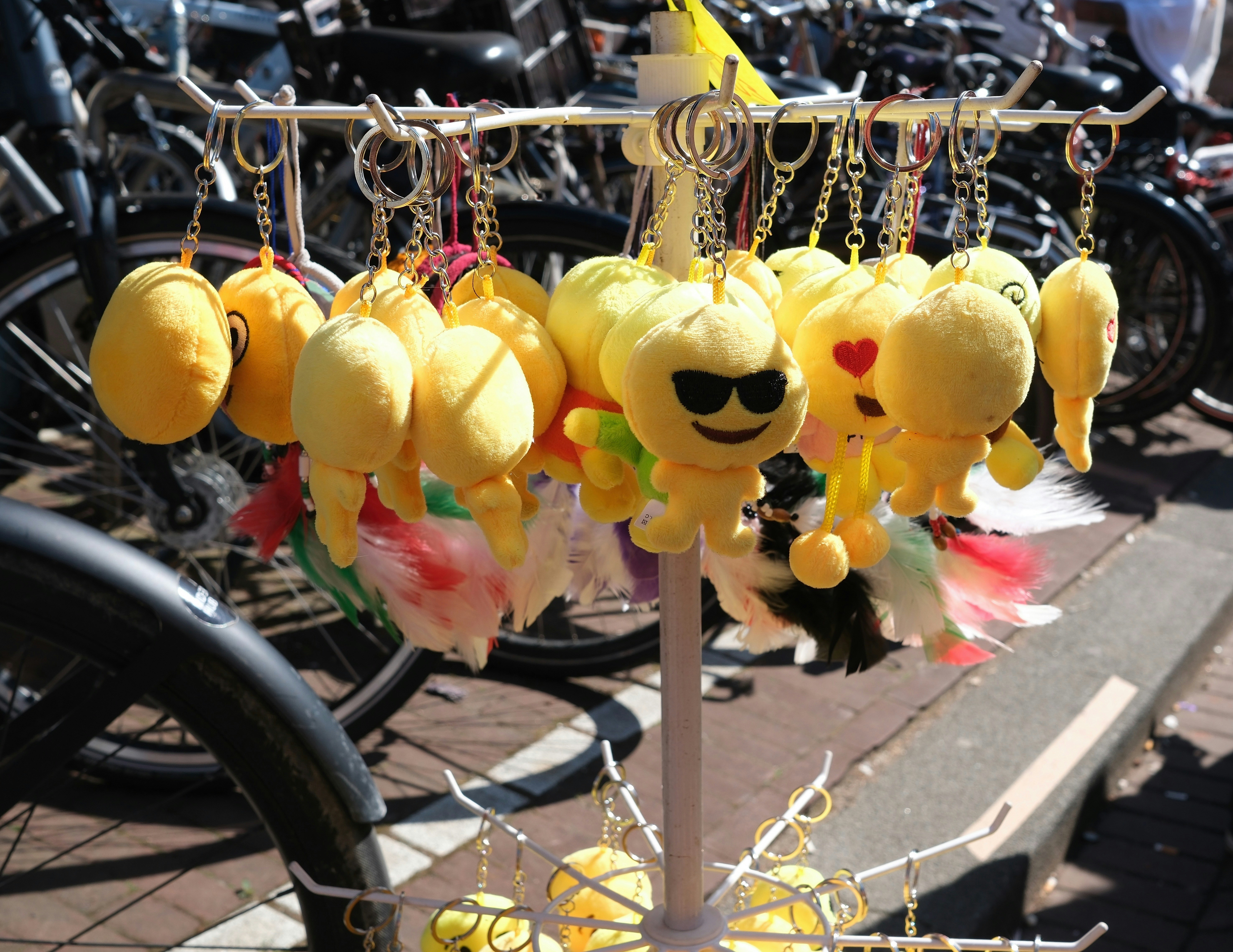 Colorful plush keychains featuring various emoji designs hang from a display stand, surrounded by bicycles in the background.