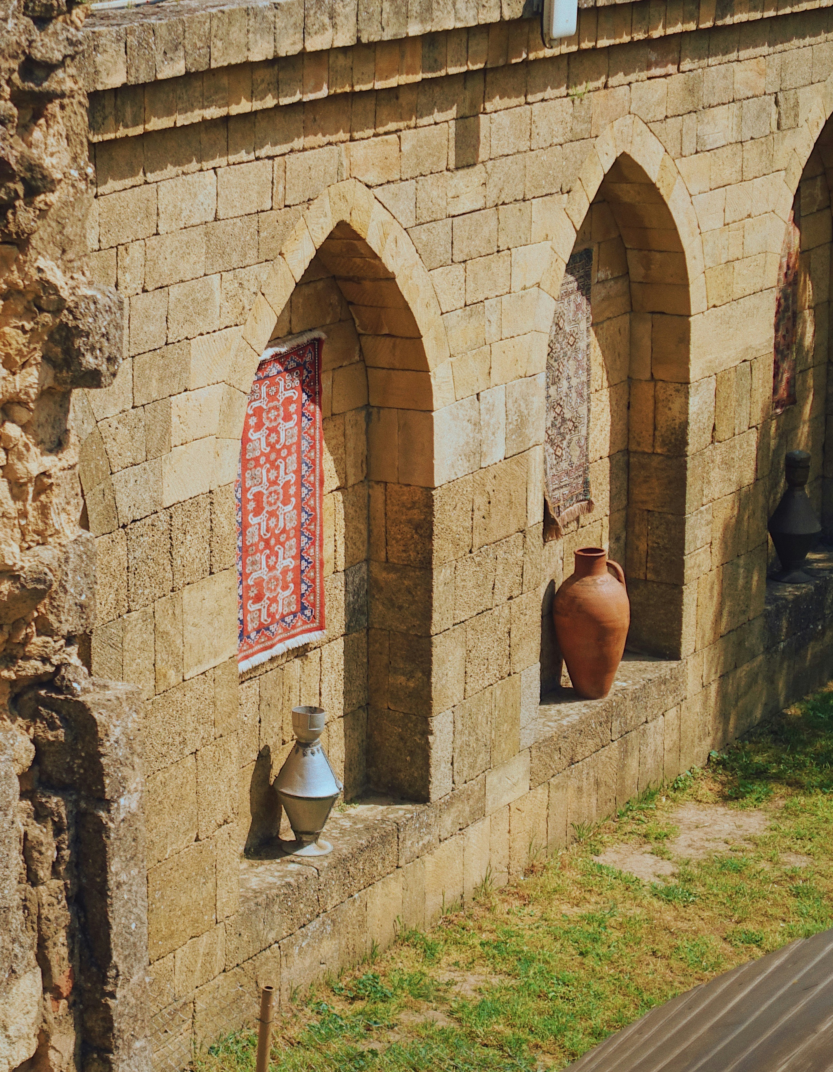 Ancient stone wall adorned with arched windows featuring intricate textiles, complemented by traditional pottery and a lush green foreground.