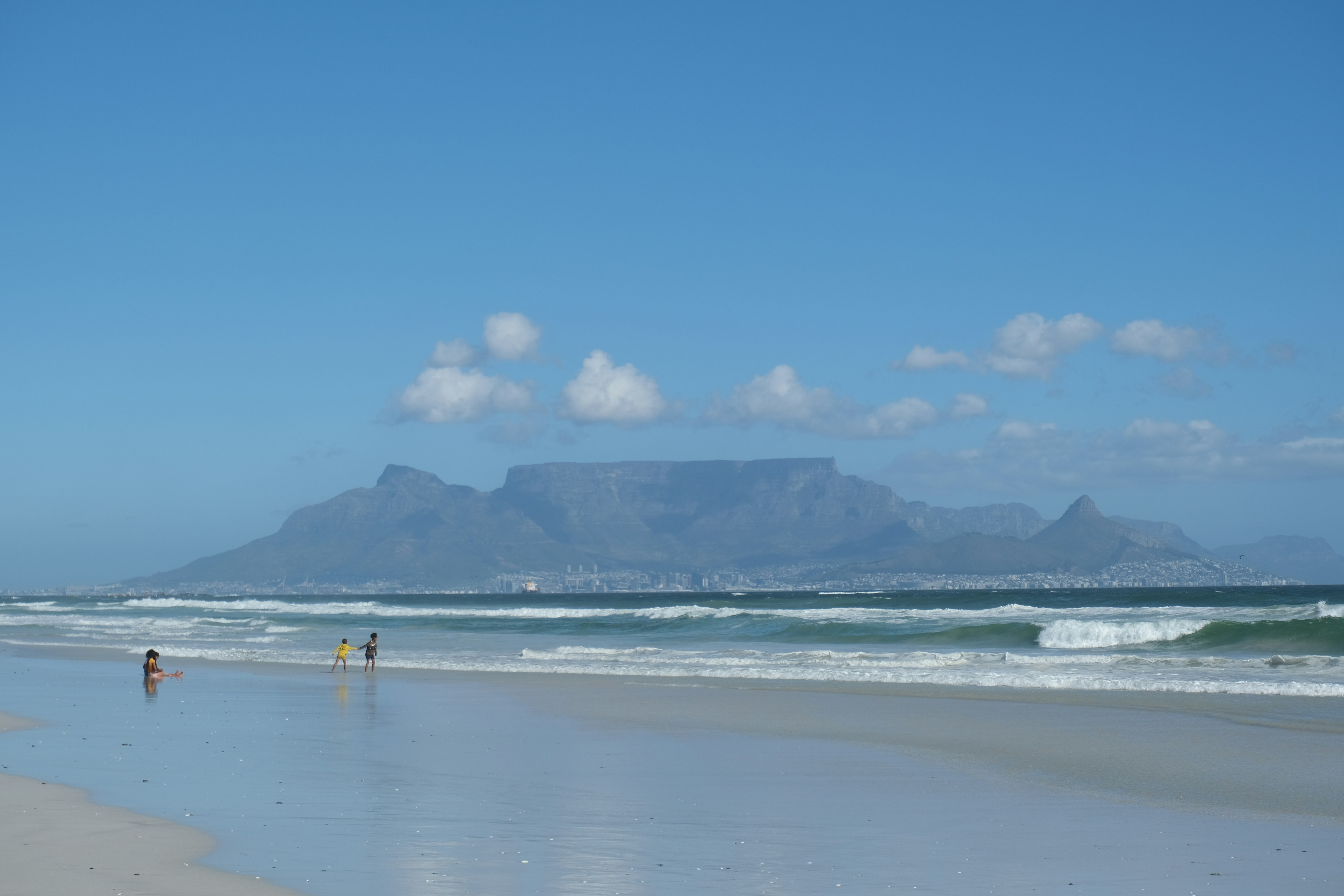 Two figures stroll along the sandy beach, with the iconic Table Mountain rising majestically in the background under a clear blue sky.