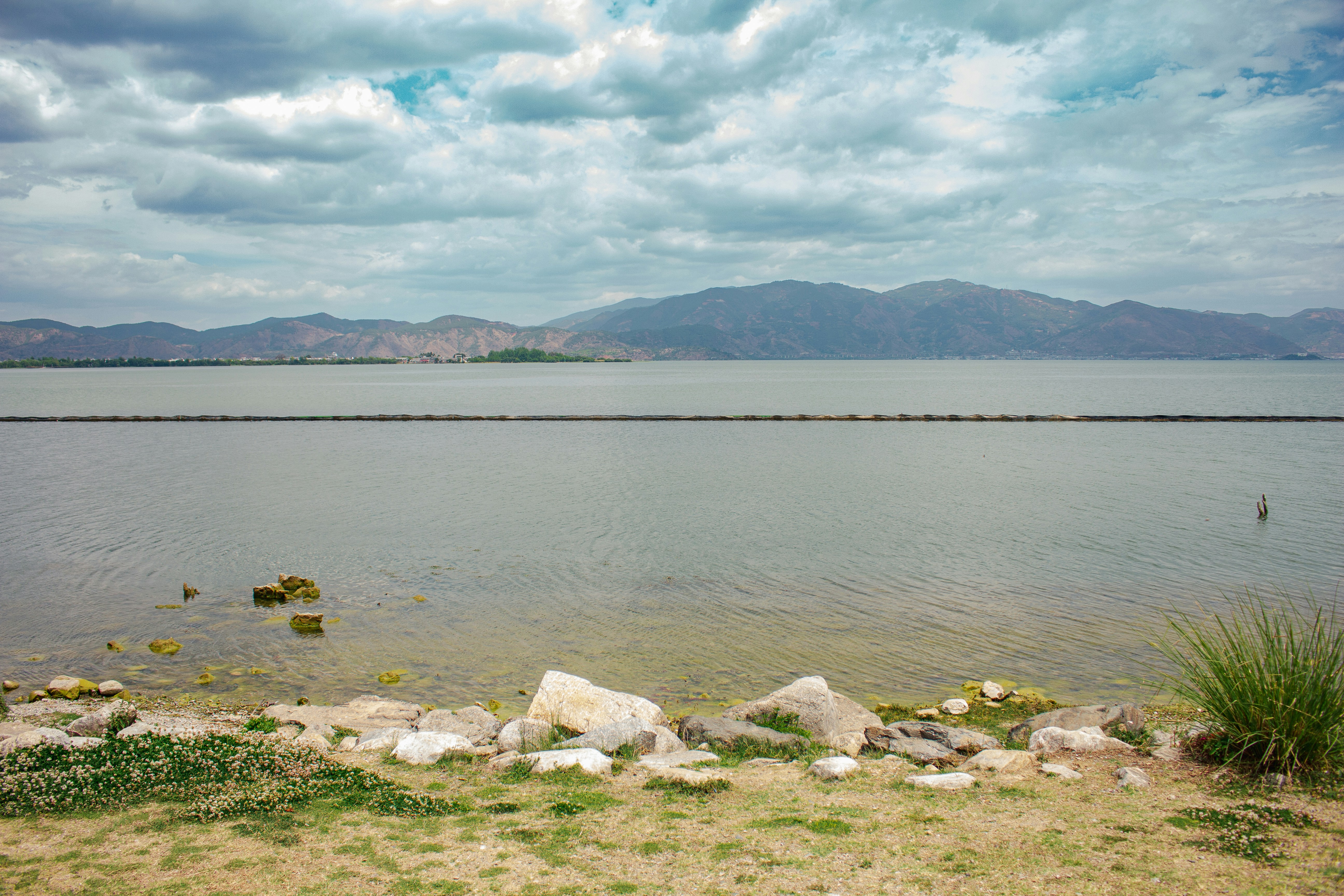 Calm lake scene with gentle waves lapping against rocky shore, framed by distant mountains under a cloudy sky.
