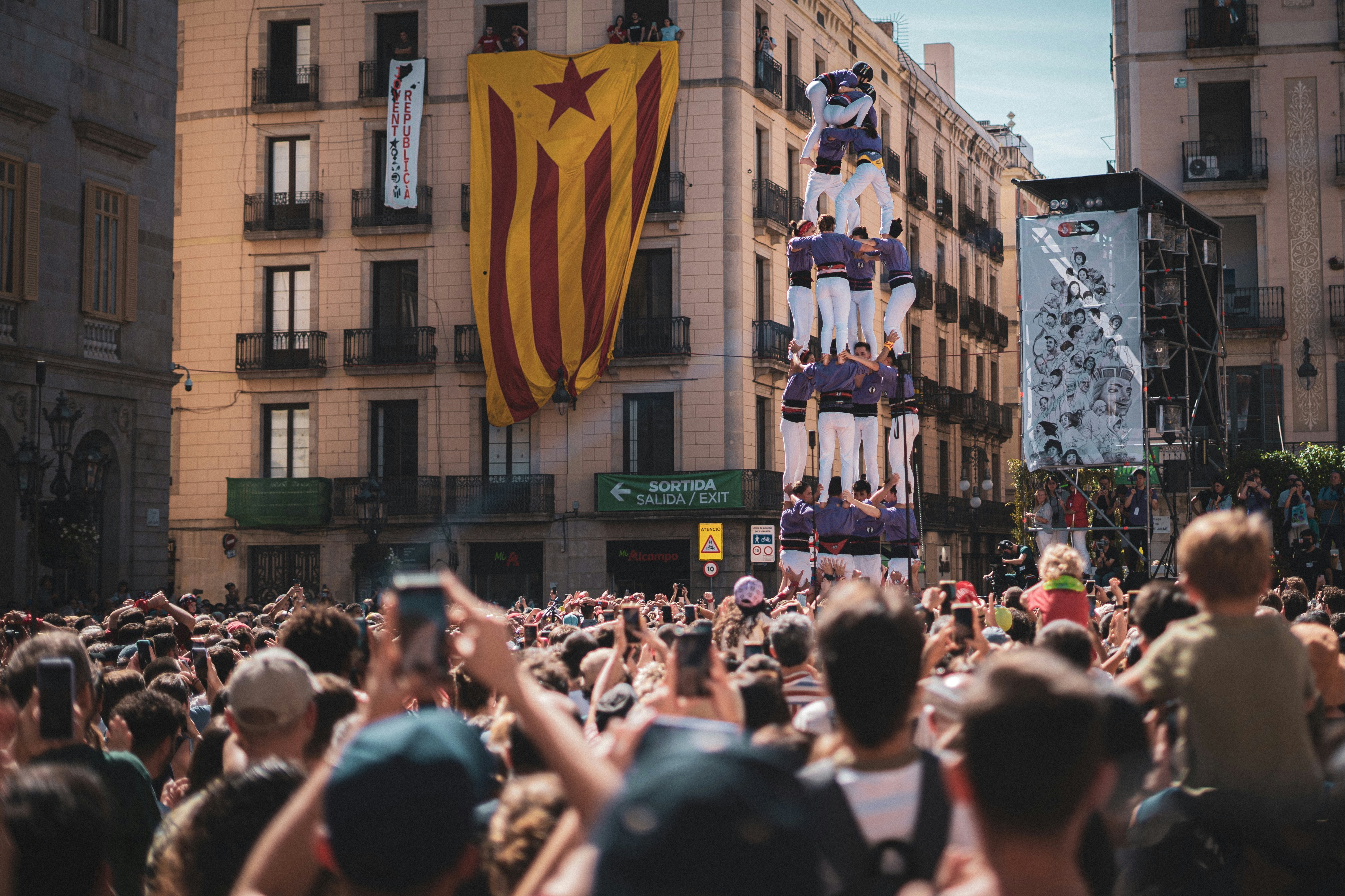 A traditional human tower, known as a “castell”, is being built by a team of castellers during a cultural festival in a bustling square in Catalonia, Spain. The scene is vibrant with spectators capturing the moment on their phones, while a large Catalan independence flag hangs from a nearby building, emphasizing regional pride and cultural heritage.