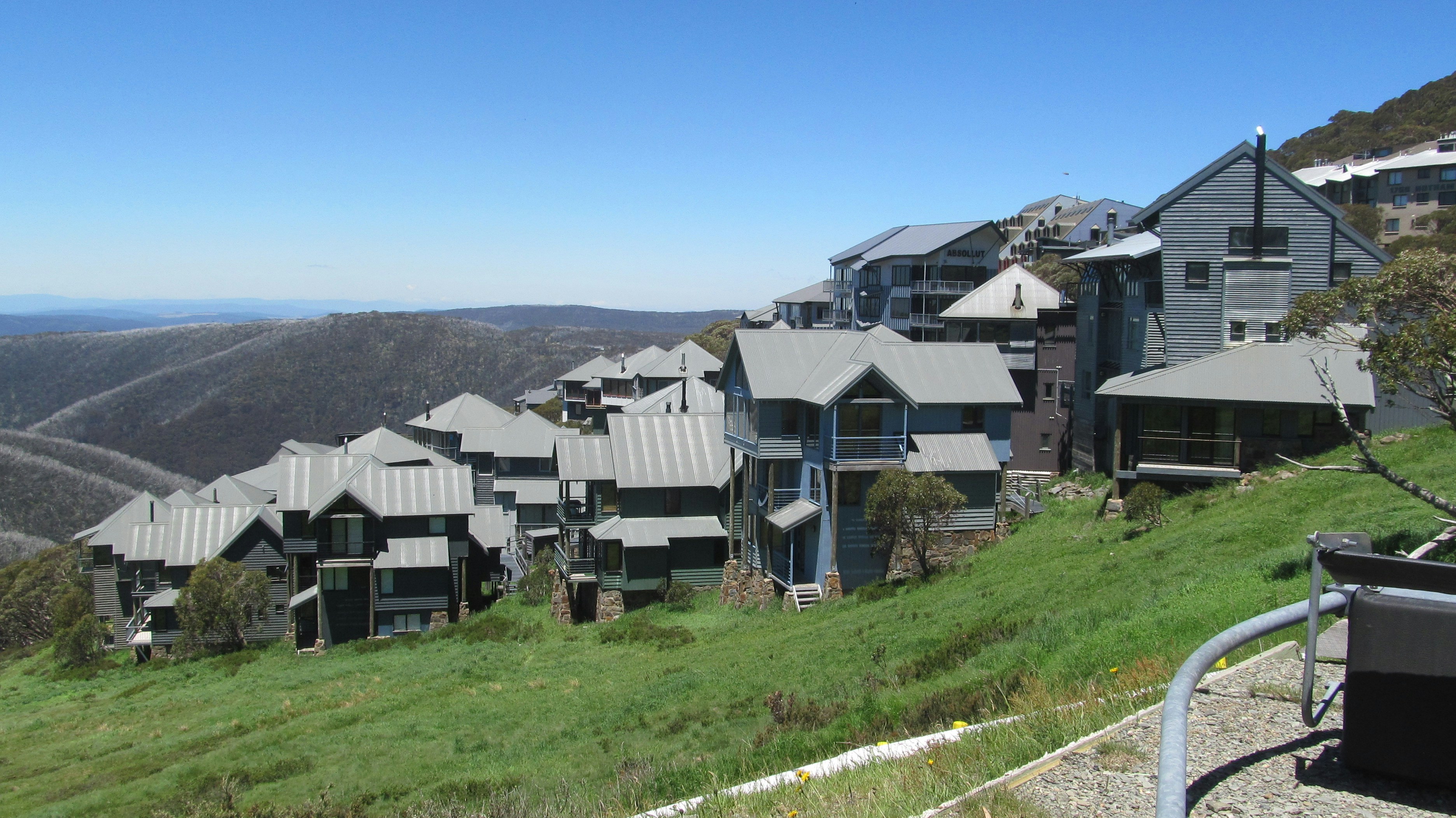 Cluster of gray, angular lodges perched on a grassy hillside with a railing in the foreground and distant mountains under a blue sky.