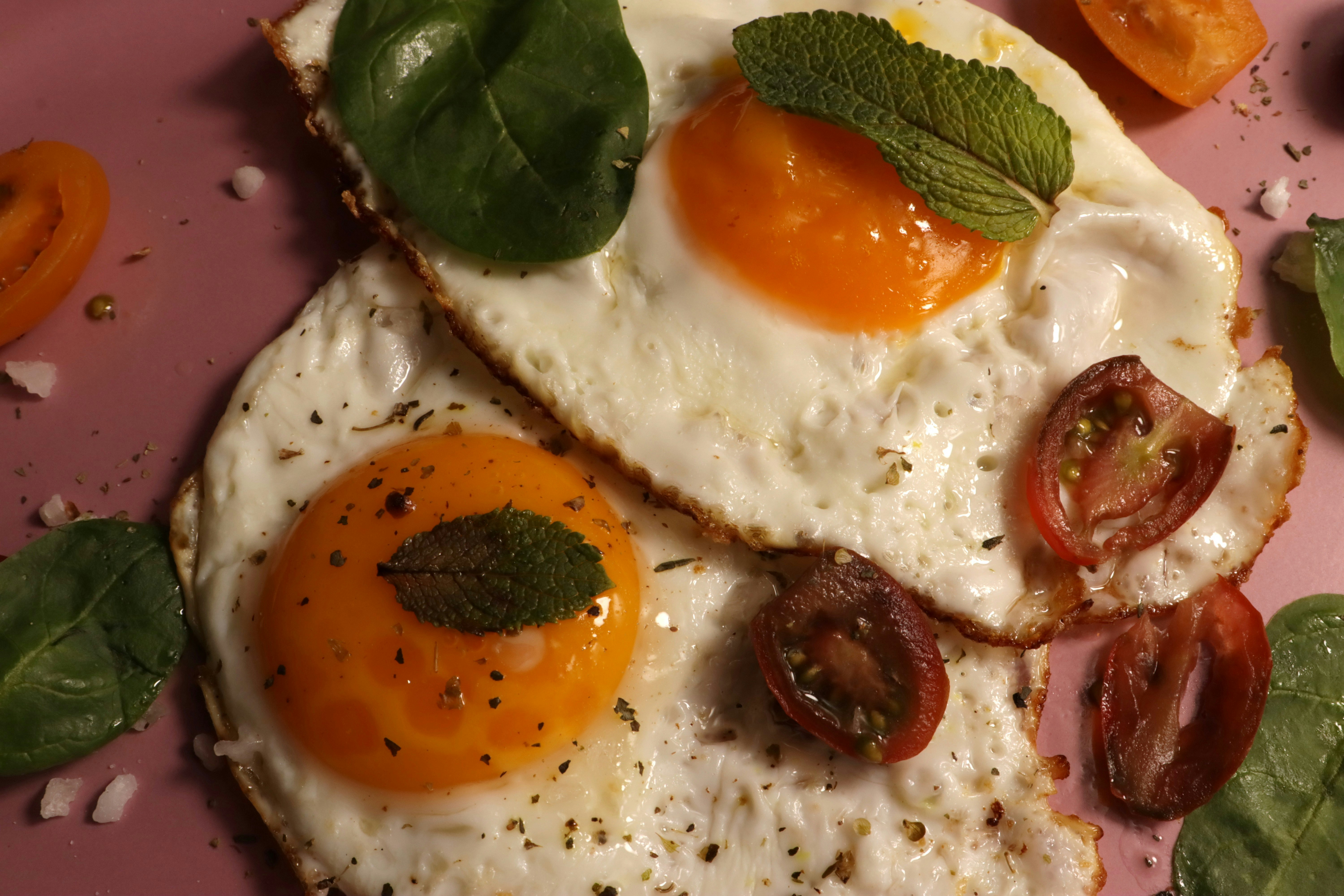 Two perfectly fried eggs garnished with fresh mint and cherry tomatoes, served on a soft pink plate.