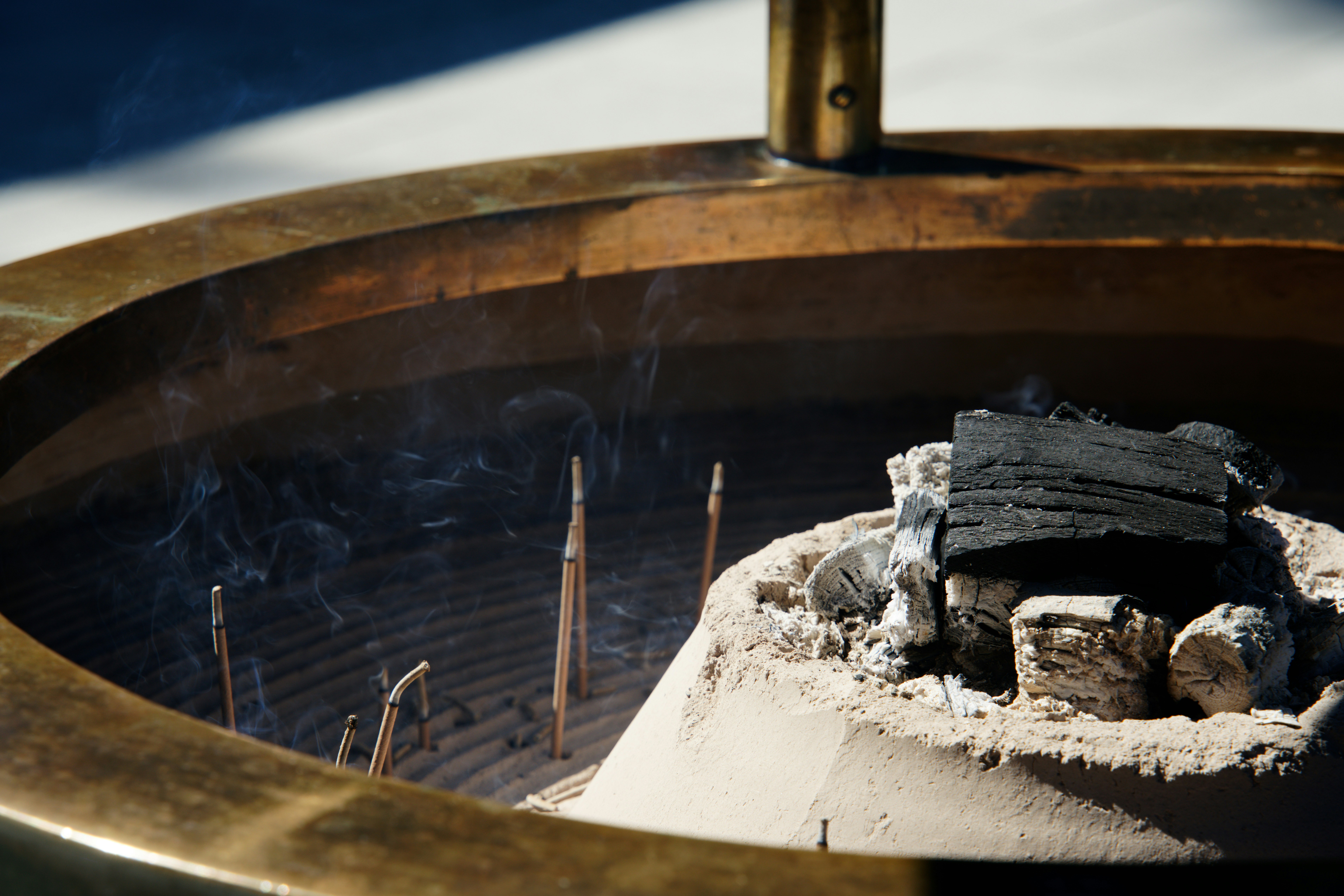 A close-up view of glowing embers surrounded by ash and incense sticks in a ceremonial bowl. The smoke gently rises, creating an atmosphere of tranquility.