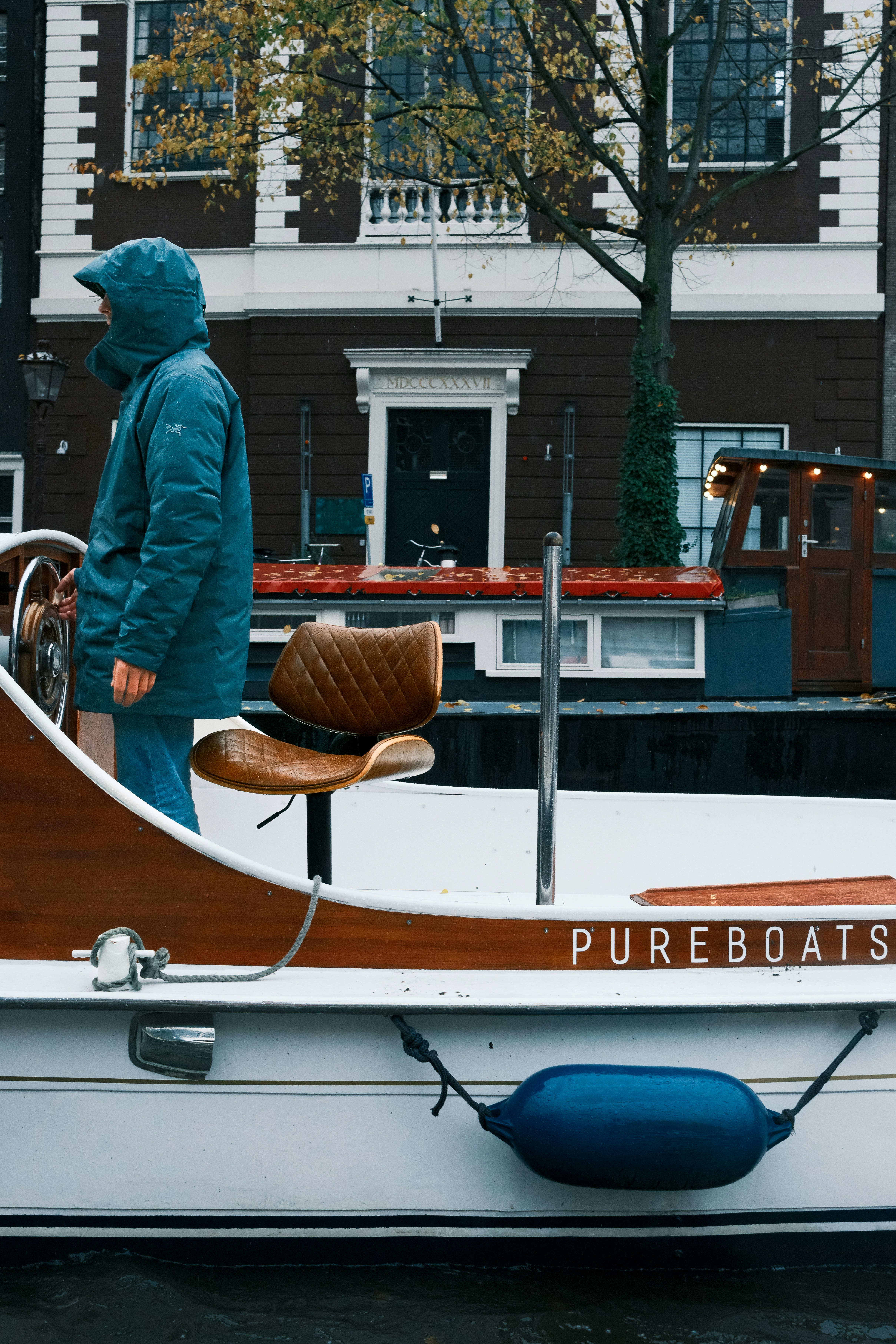 Blue-hooded silhouette on a canal boat beside a brick townhouse in autumn light