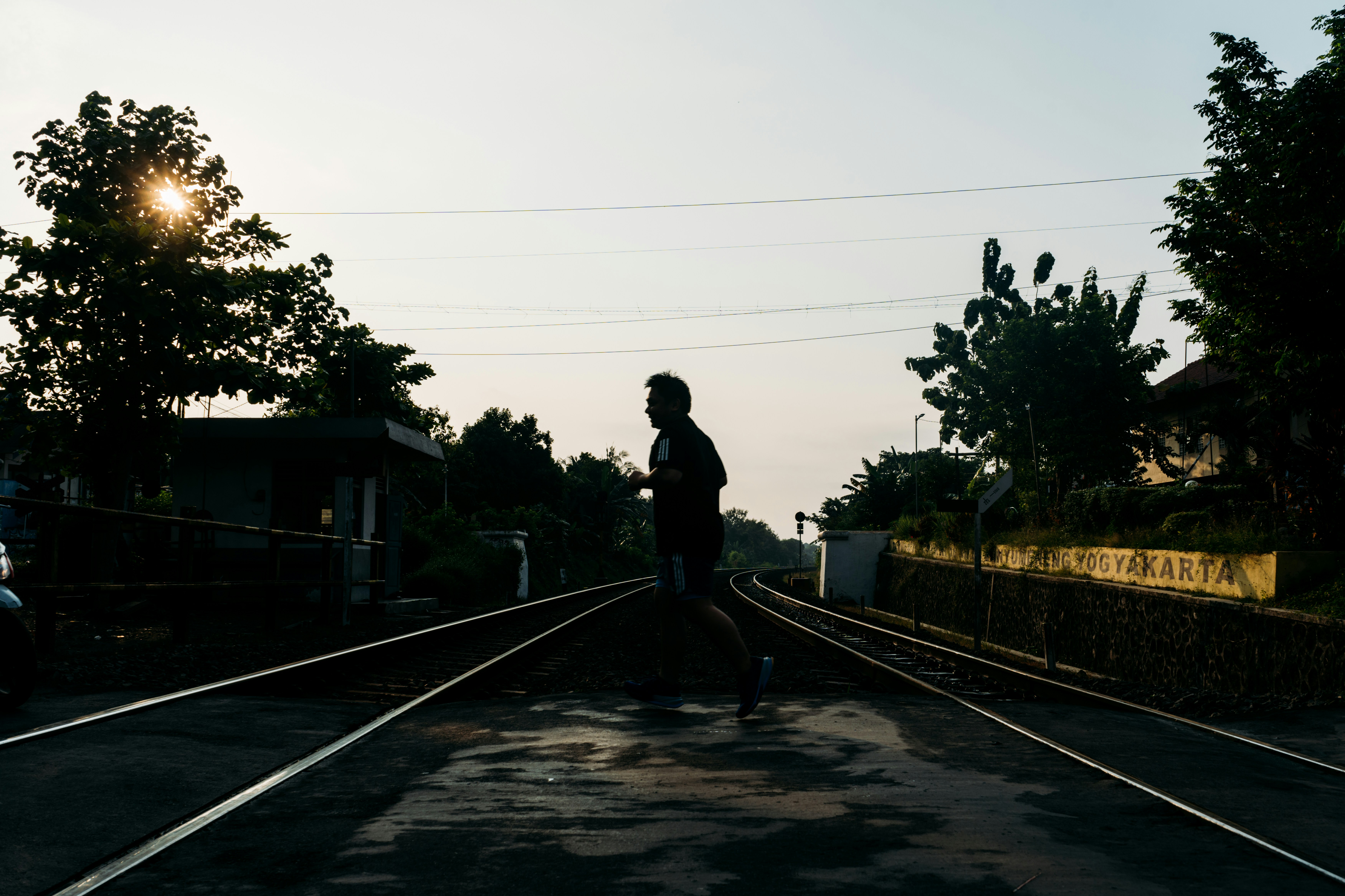 Silhouette of a runner crossing railway tracks during sunrise, framed by lush greenery and urban elements.