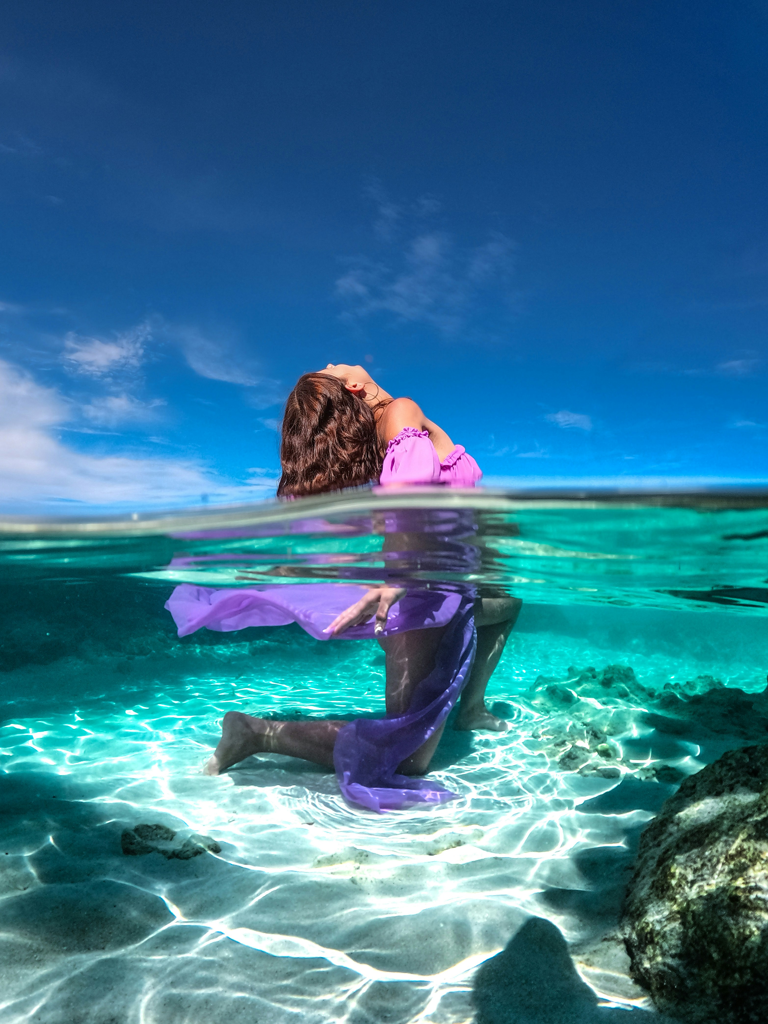 Split-View Serenity: Violet dress drifts in crystalline turquoise shallows under a bright sky