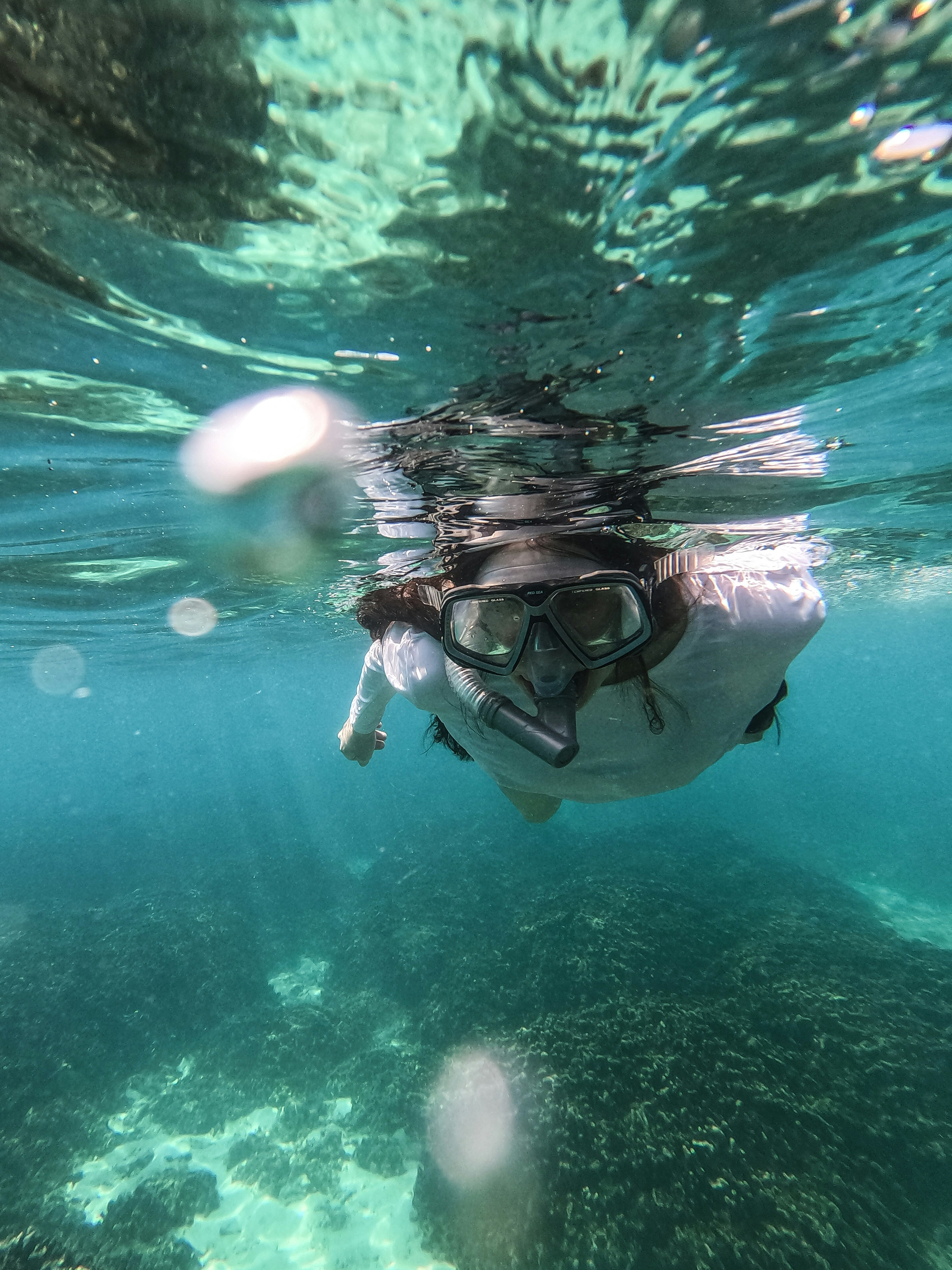 An underwater diver wearing a snorkel mask swims toward the camera, with the mask and mouthpiece clearly visible in clear turquoise water. Sunlit seabed and drifting light orbs glow beneath.