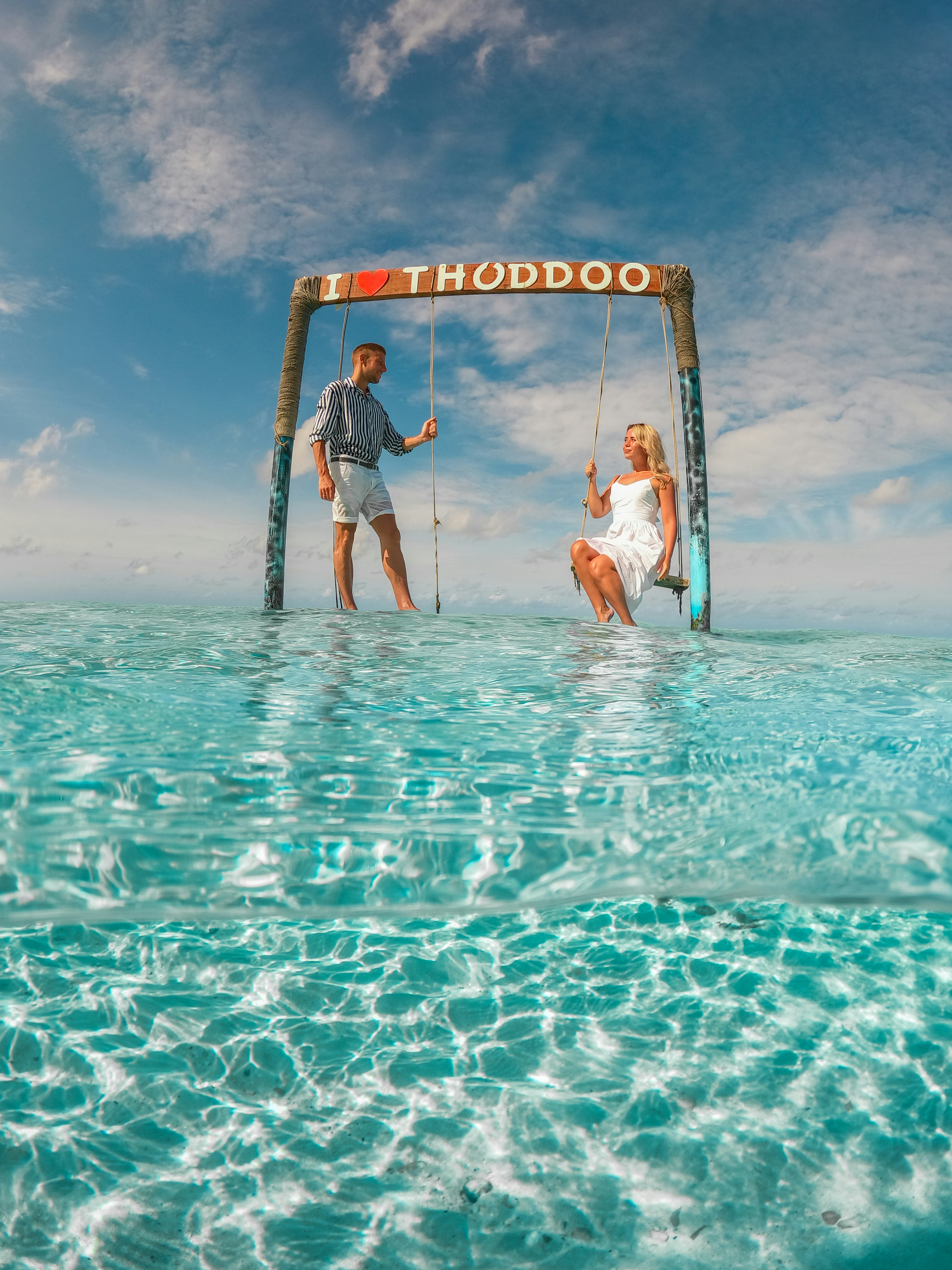 A photograph of a couple perched on an over-water swing frame above a turquoise lagoon, beneath a bright blue sky. The wooden frame reads 'I love Thoddoo'.