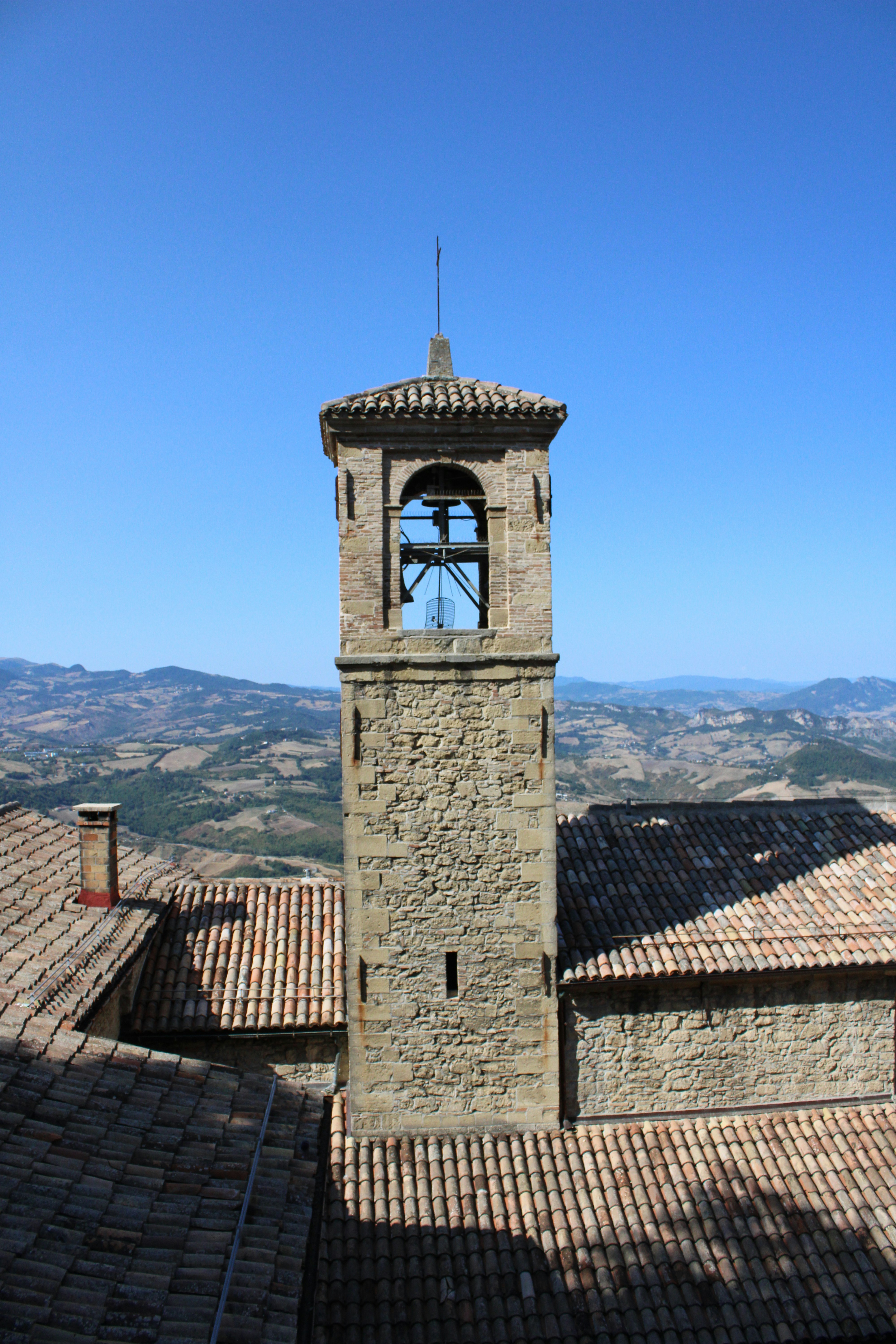 Stone Bell Tower Rising Over Terracotta Roofs, Framed by a Distant Valley