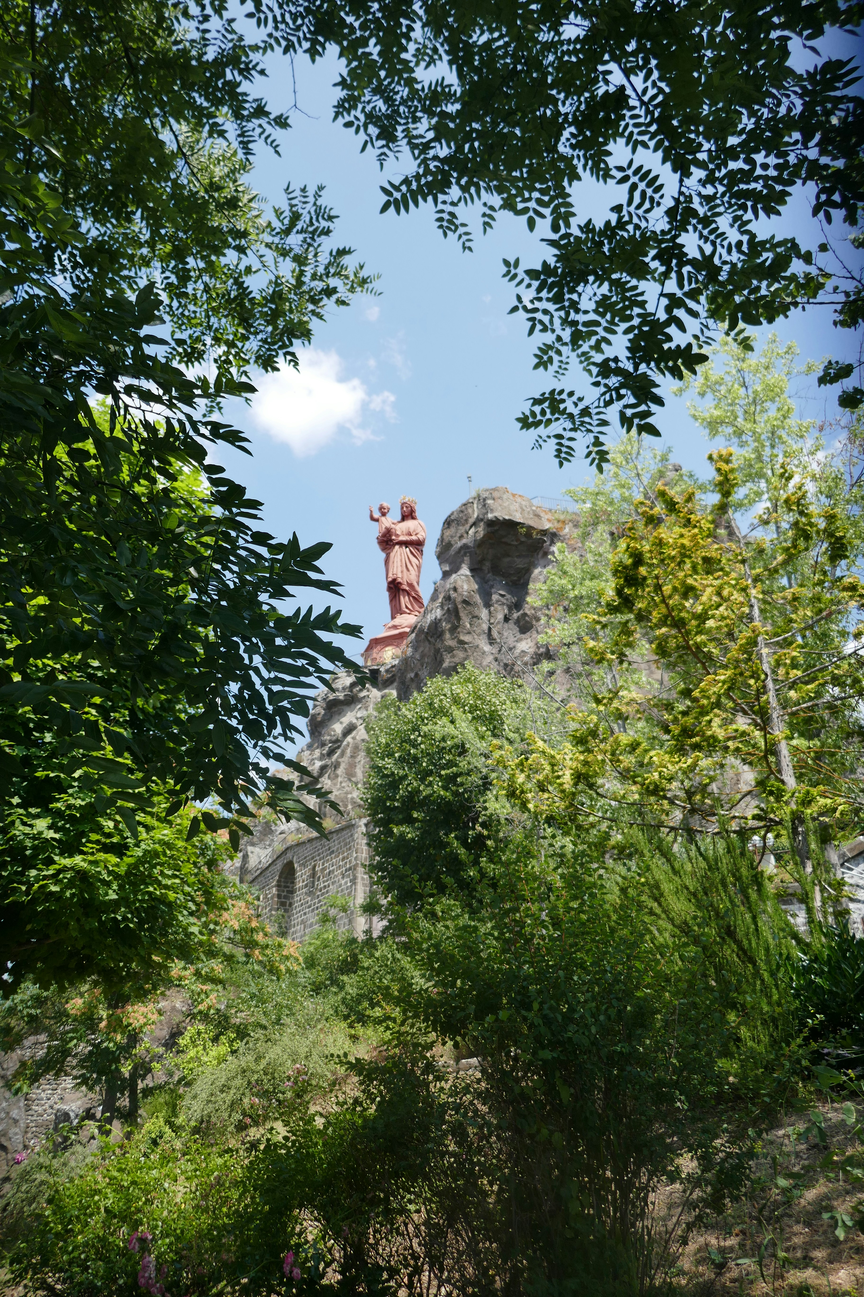 Terracotta statue atop a rocky outcrop rises above a lush garden frame as branches shelter the scene, with a bright blue sky behind.