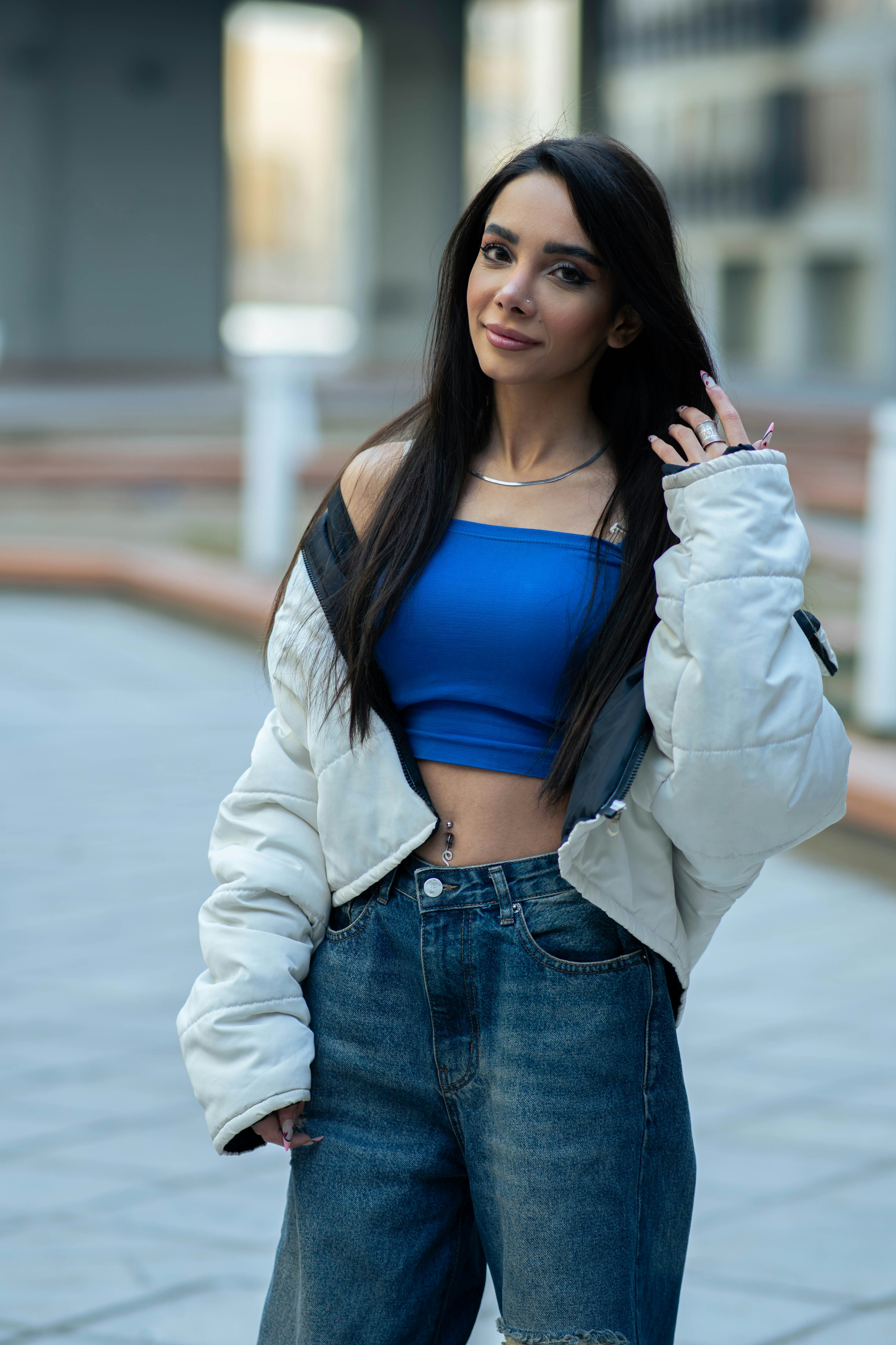 A young woman poses confidently in an urban setting, wearing a cropped blue top and oversized jacket, with a stylish flair. Her relaxed demeanor adds to the contemporary vibe.