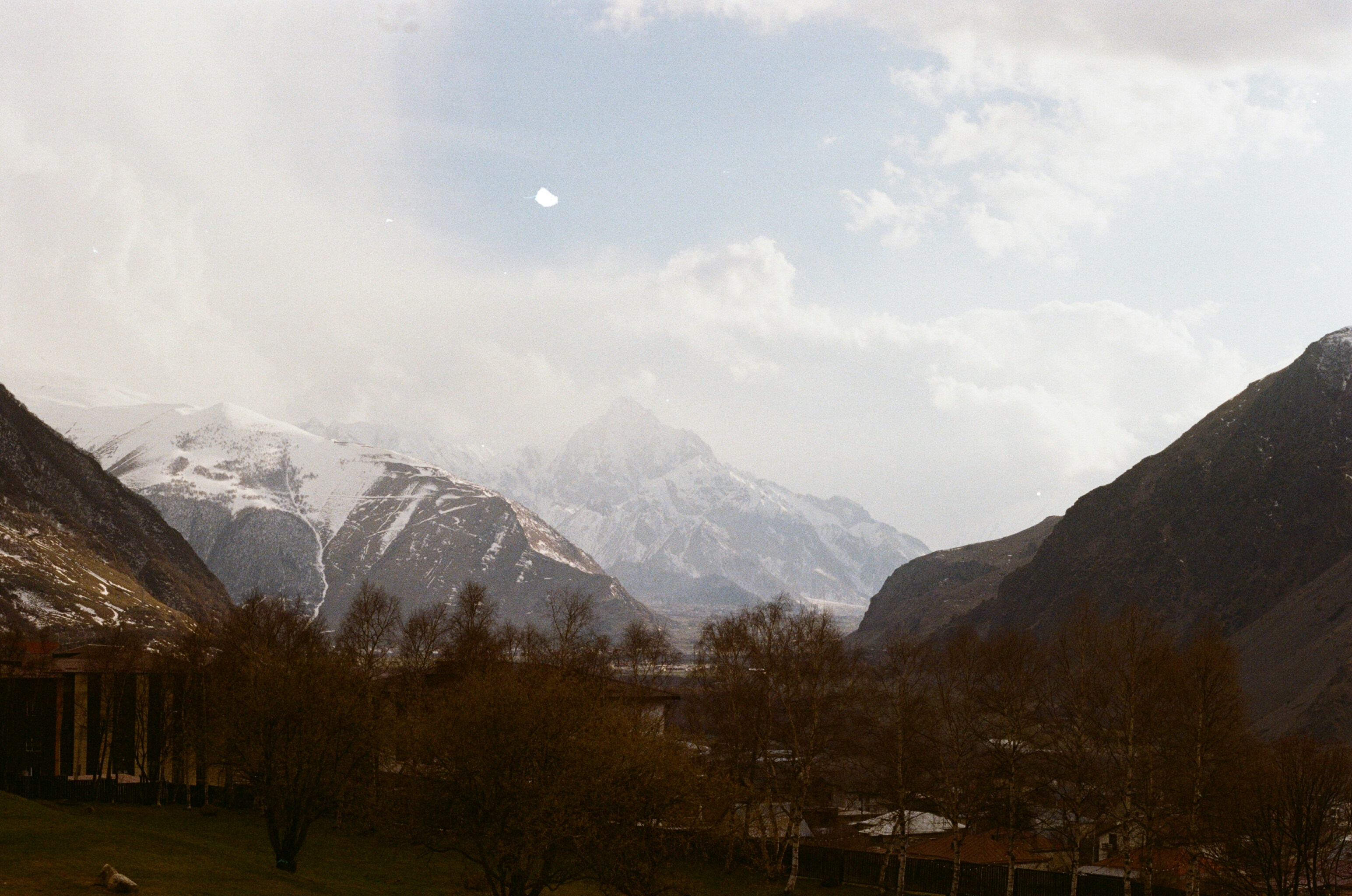 Majestic mountain range under a cloudy sky, showcasing snow-covered peaks and a lush valley below. The scene captures the serene beauty of nature's untouched landscapes.