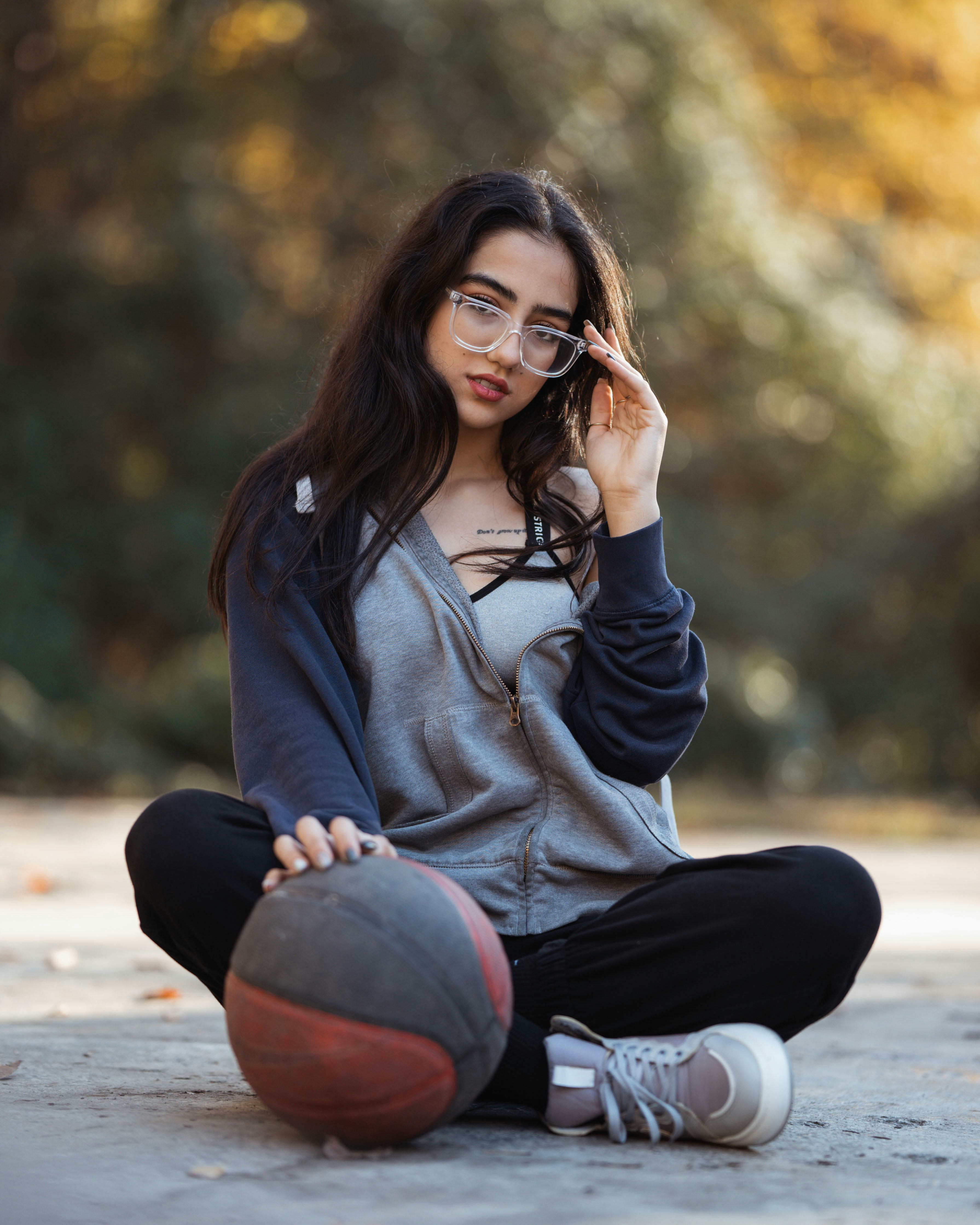 Young woman in casual attire sits cross-legged on the ground with a basketball, exuding a relaxed yet focused vibe amidst a blurred natural backdrop.