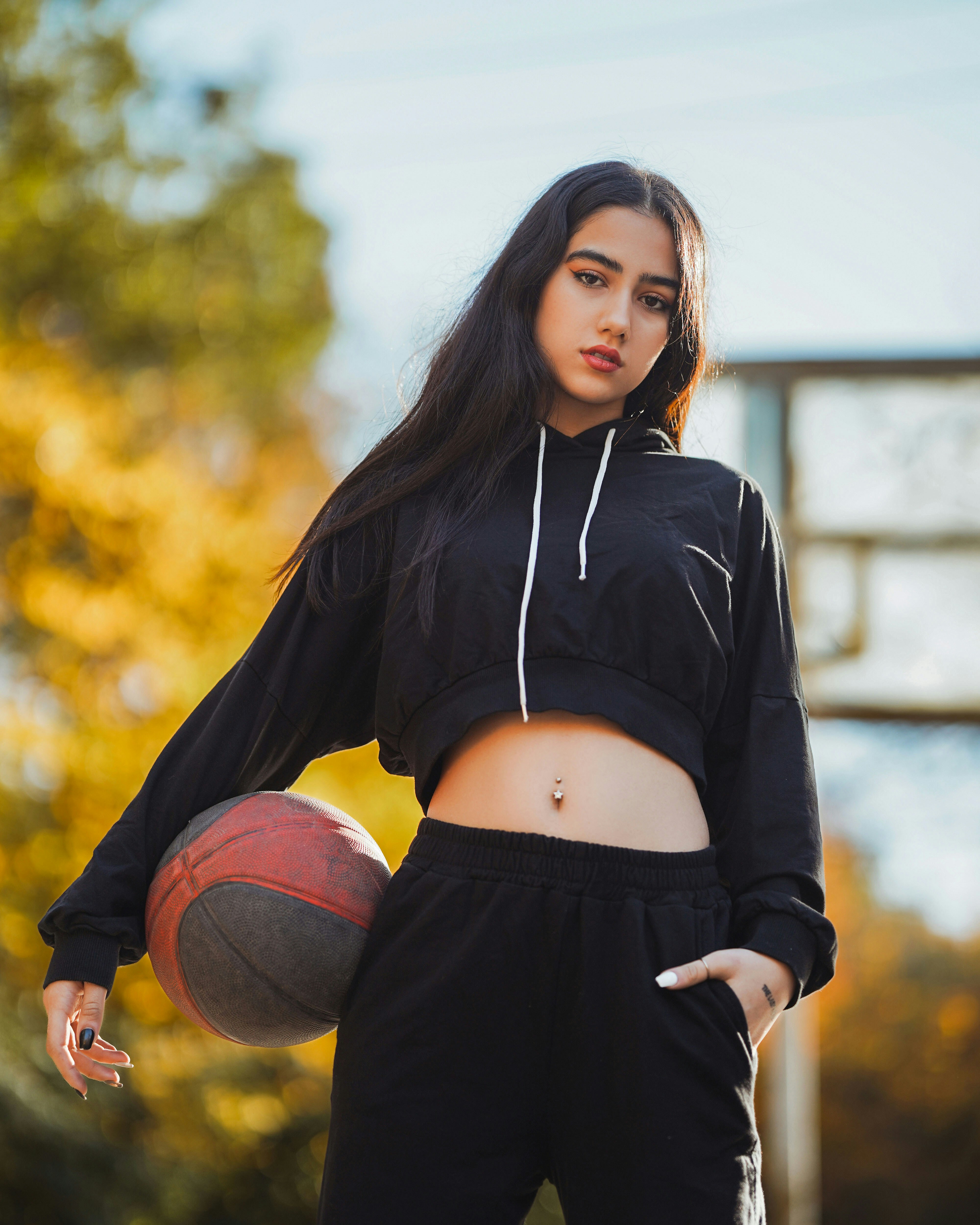 Young woman in a cropped hoodie and sweatpants holds a basketball, exuding confidence against a backdrop of autumn foliage.