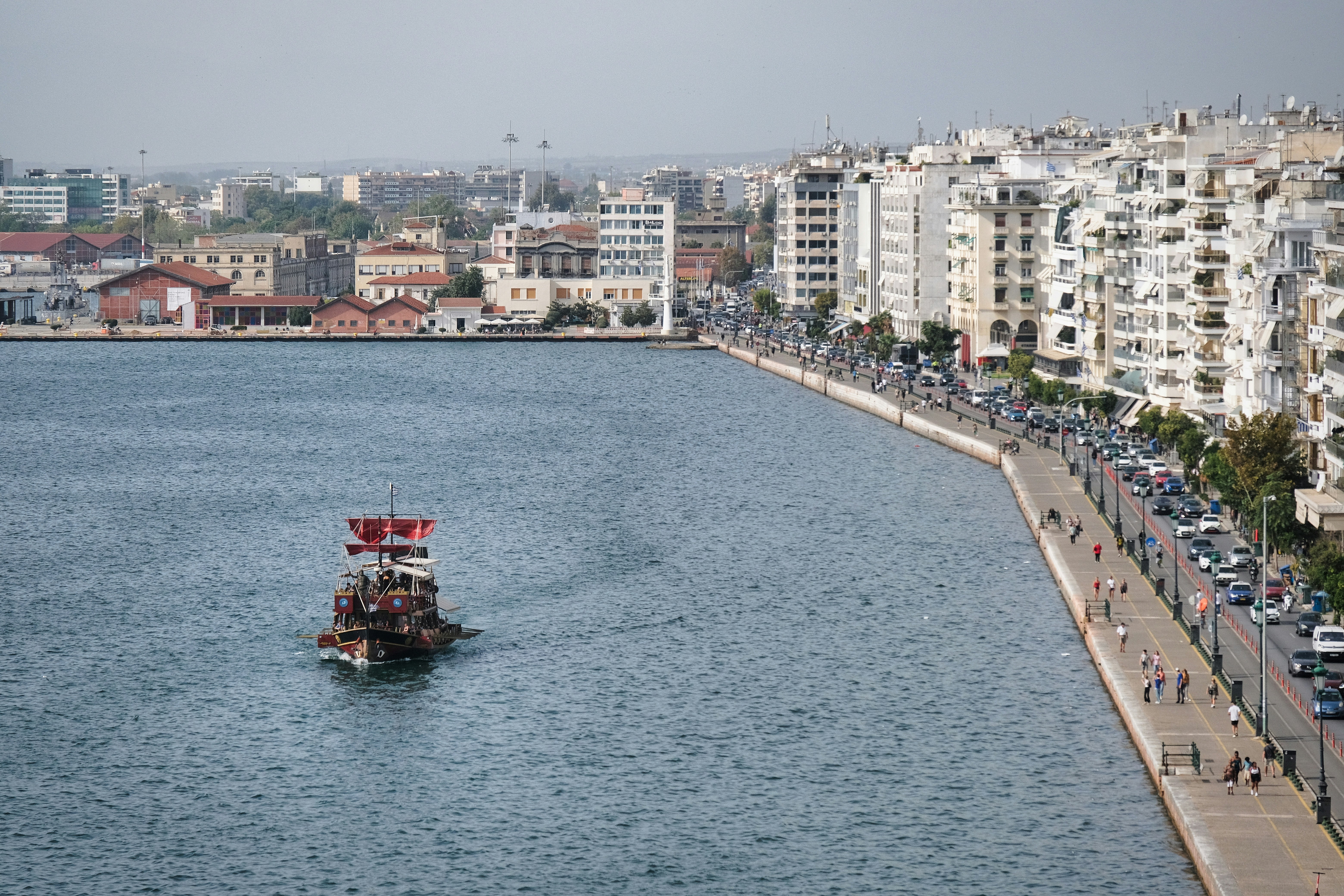 A vibrant seaside moment where the blue waters of the Thermaic Gulf meet the elegant urban shoreline. A boat styled like a pirate ship, with red canopies and a festive atmosphere, moves through the frame from the left, bringing dynamism to the scene. To the right, the waterfront promenade bustles with walkers, joggers, and parked cars as modern apartments and historical buildings stand in tight succession, showcasing the city’s Mediterranean charm. The contrast between the relaxed movement on the sea and the structured, busy life on land tells a layered story of Thessaloniki's character.