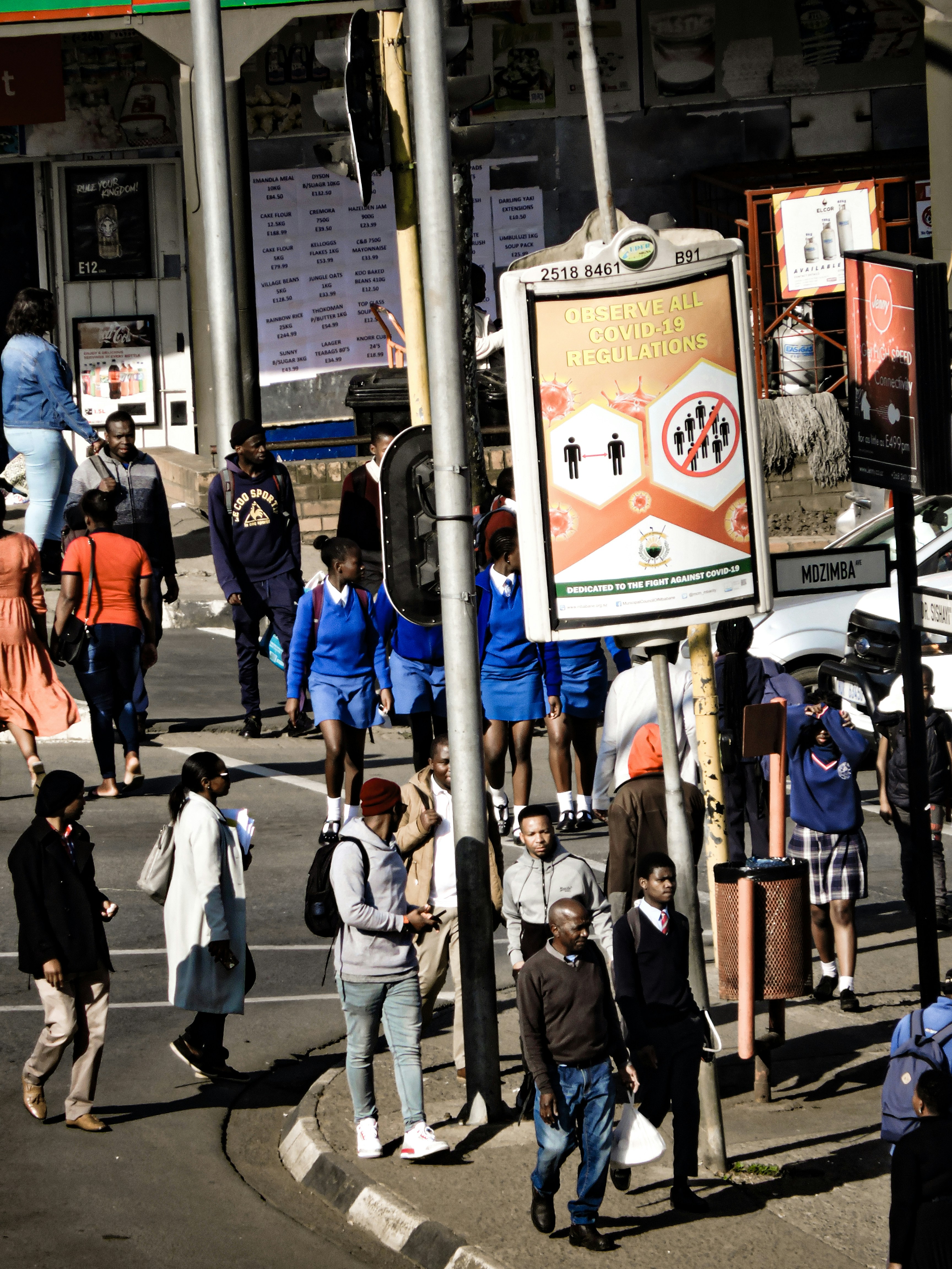 Crowded urban crosswalk with pedestrians of varied attire, including a cluster in blue uniforms, beside a prominent COVID-19 regulations poster at a street corner.