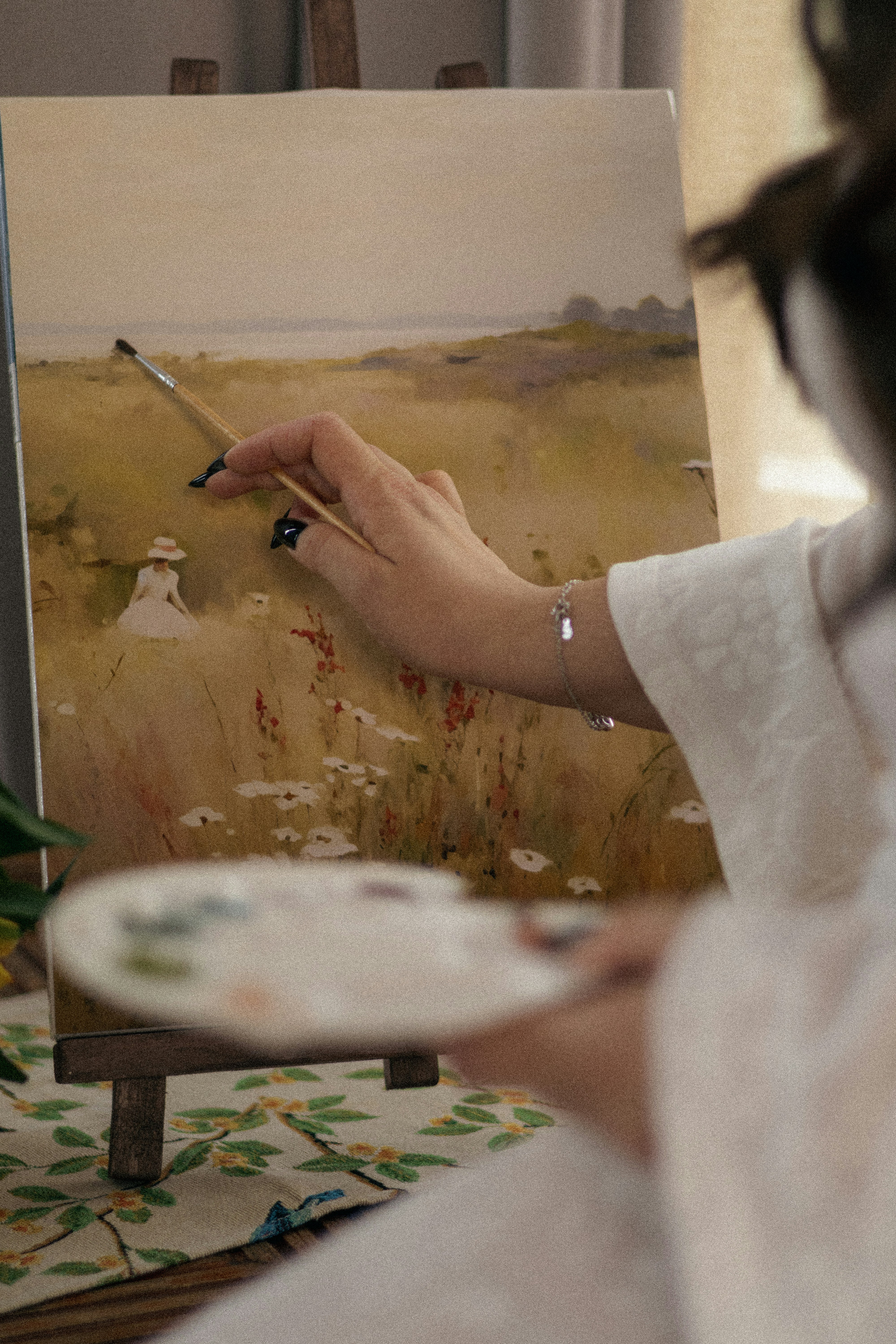 A painter's hand guides a brush across a landscape painting on an easel, with a paint palette visible in the foreground.