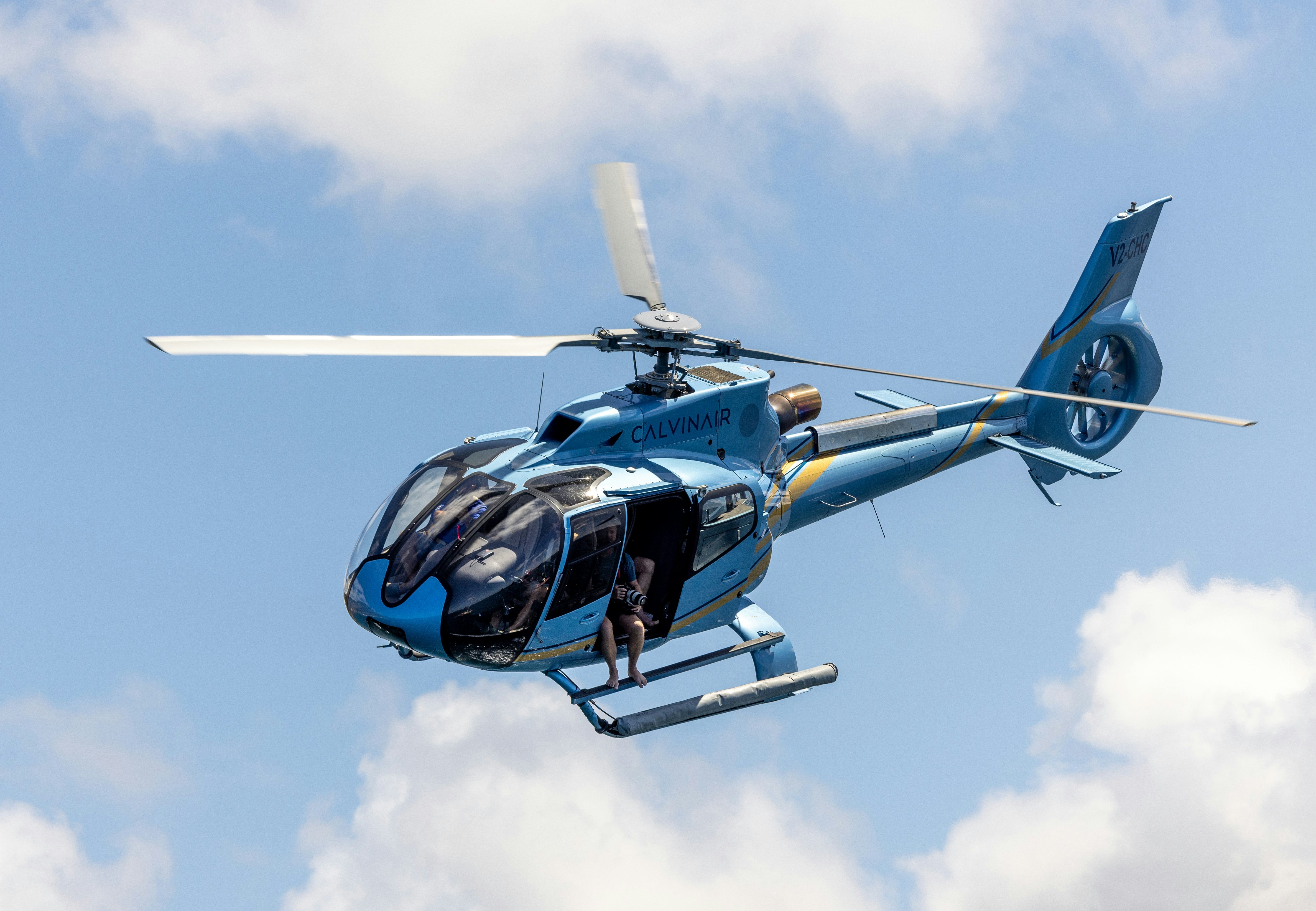 A blue helicopter in flight: cockpit and passenger visible against a sky of white clouds