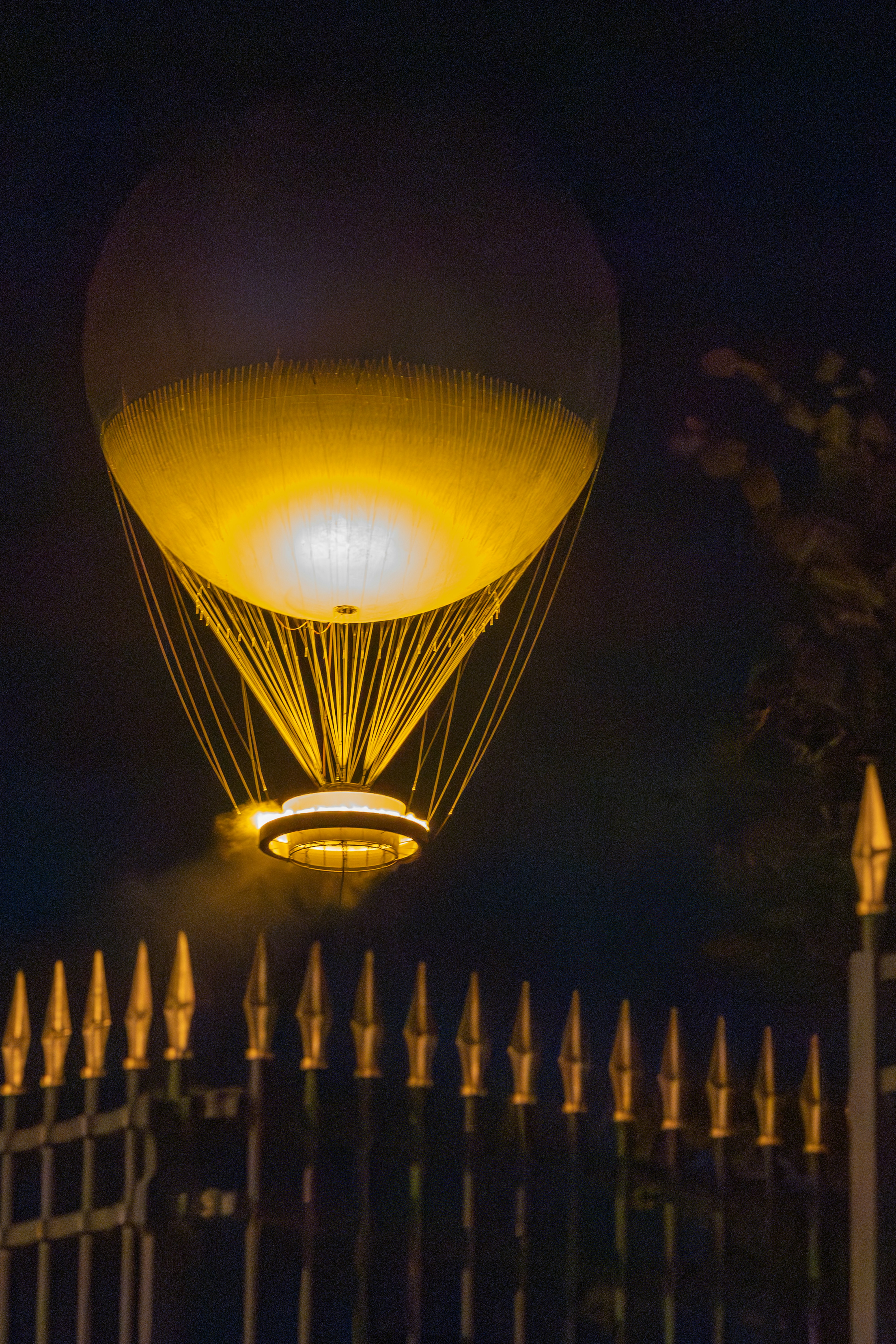 A glowing hot air balloon hovers above a decorative fence at night, casting a warm light in the darkness. The scene captures the enchanting moment of flight.