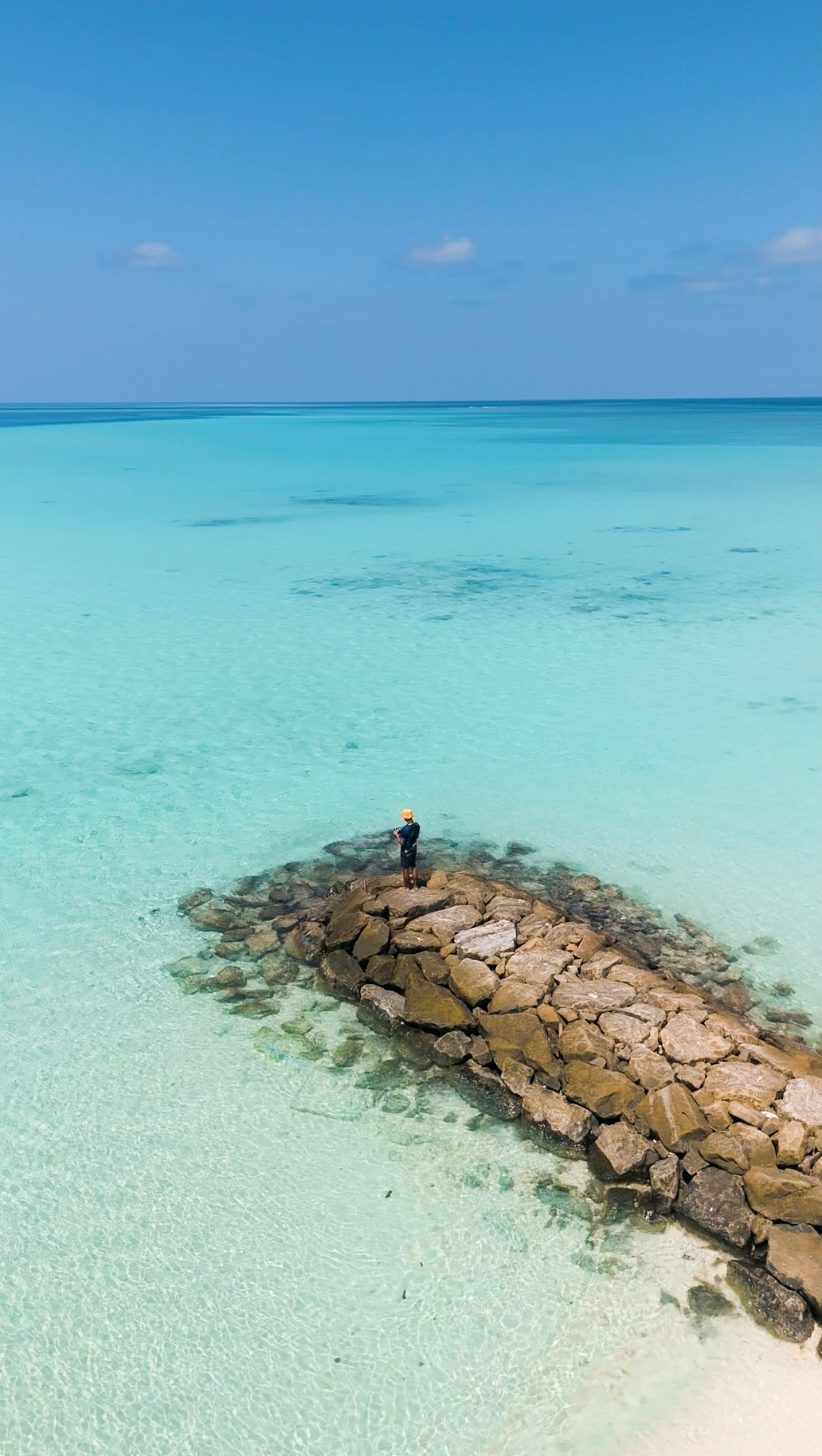 A lone figure stands on a rocky jetty, surrounded by crystal-clear turquoise waters under a bright blue sky.