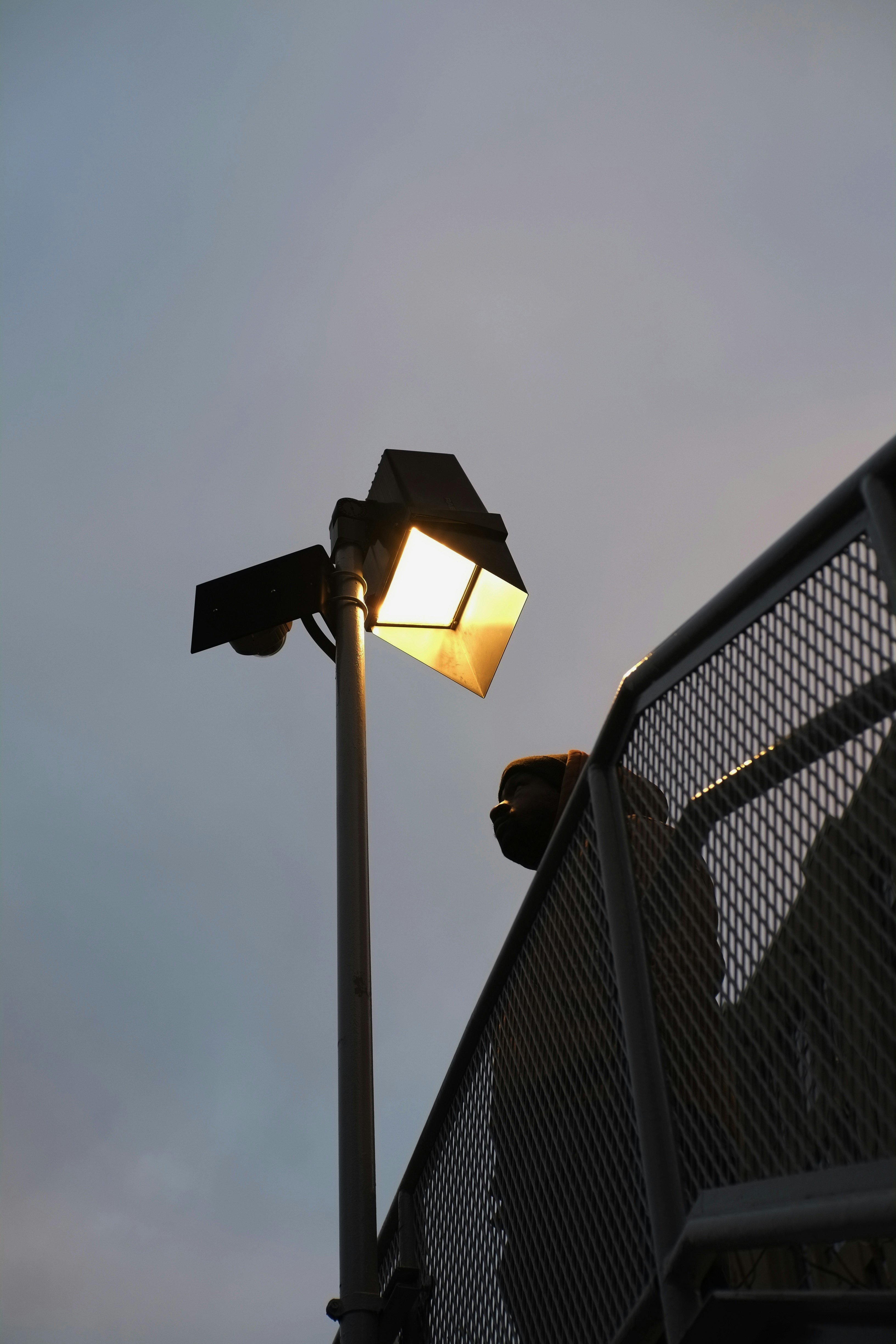 A streetlight casts a warm glow against a moody sky, highlighting the geometric lines of a nearby railing. The scene evokes a sense of urban tranquility.