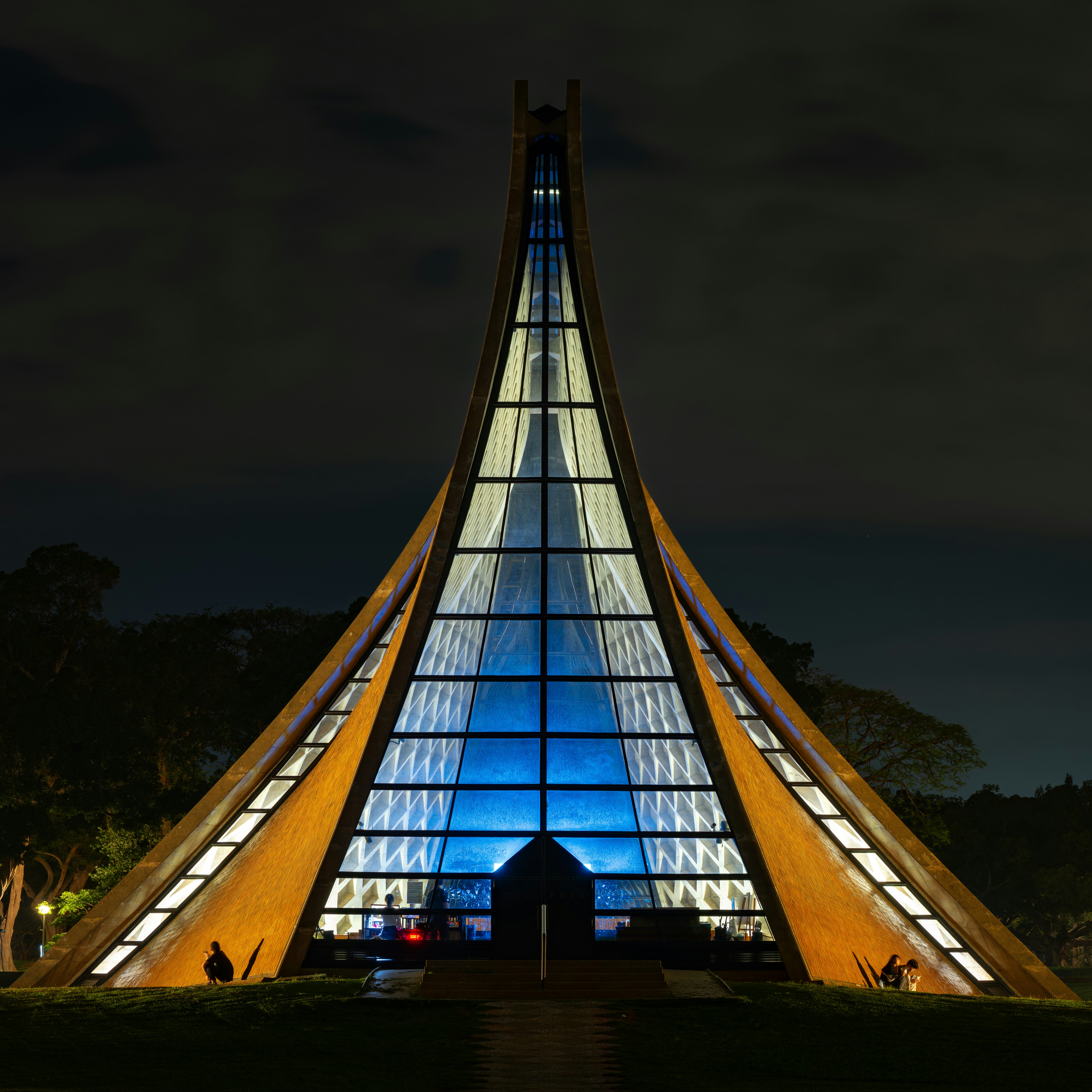 A striking modern chapel with a soaring glass facade illuminated by blue lights, set against a dark sky. The unique triangular design draws the eye upward.