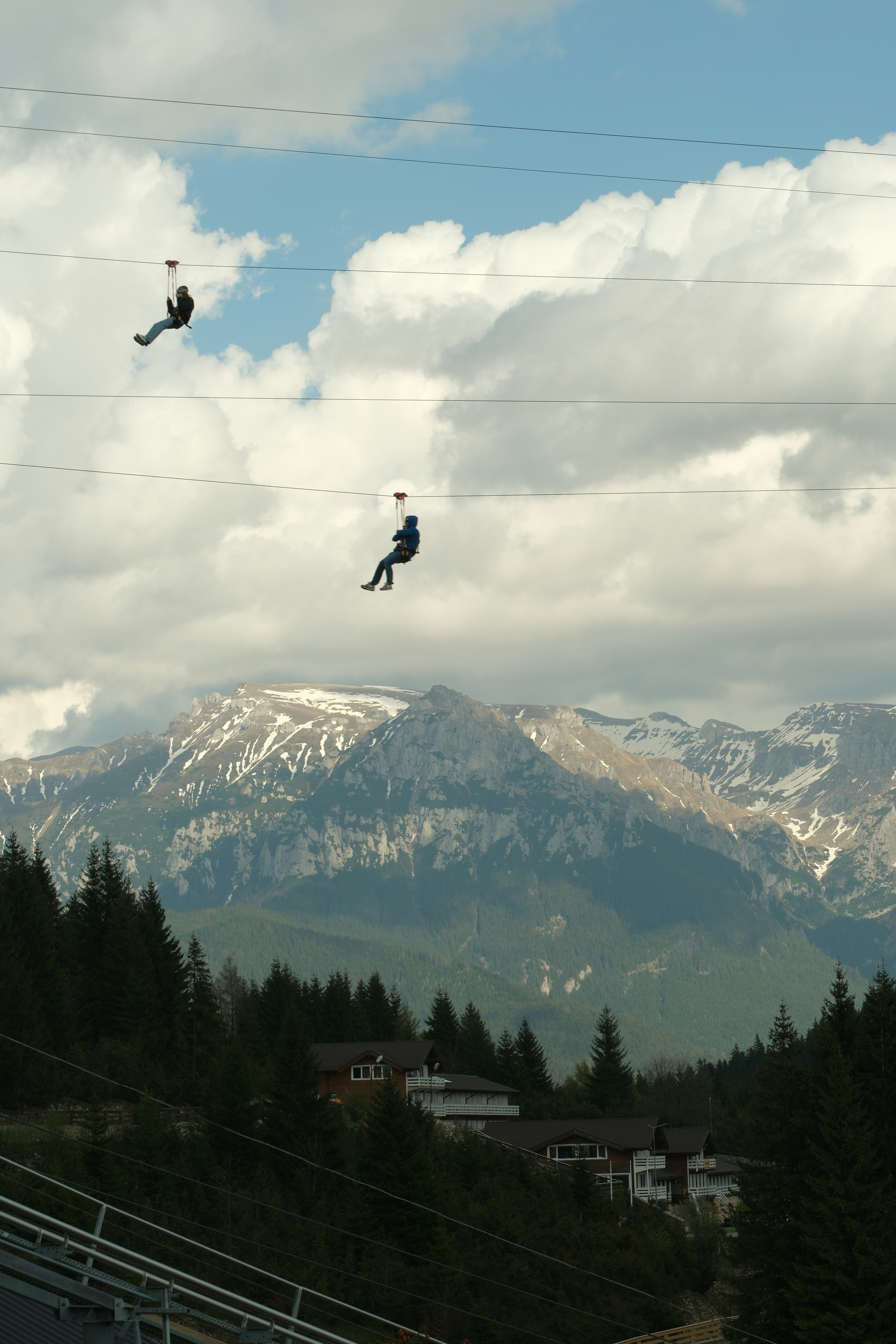Two zipliners glide above a scenic landscape featuring mountains and a forested area. The dramatic clouds add depth to the sky.