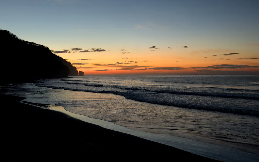 Sunset-lit coastline with a dark cliff silhouette on the left and gentle waves reflecting an orange-to-blue sky.