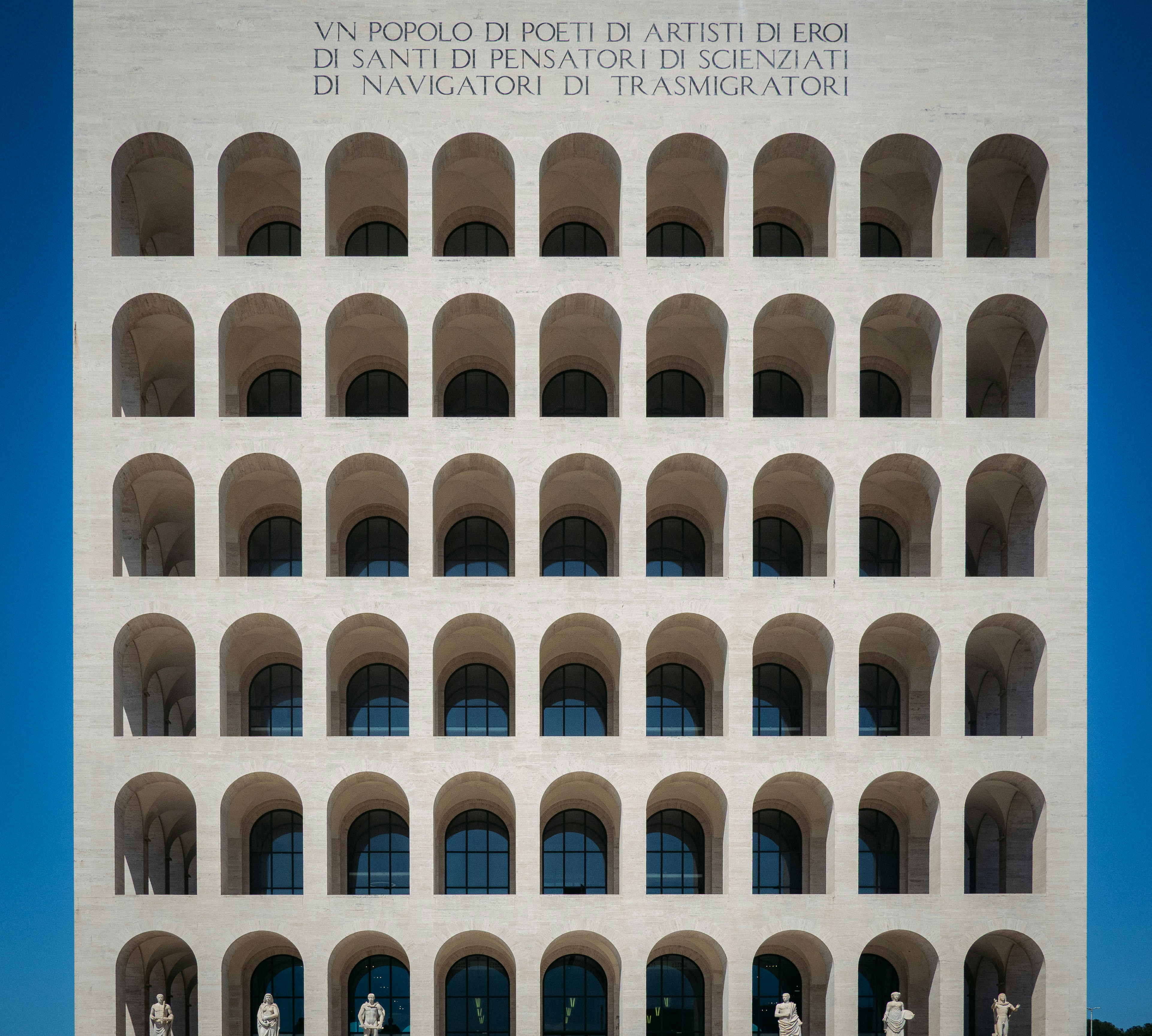 Symmetrical building facade featuring rows of rounded arches in light stone, framed by blue borders and topped by an inscribed band.