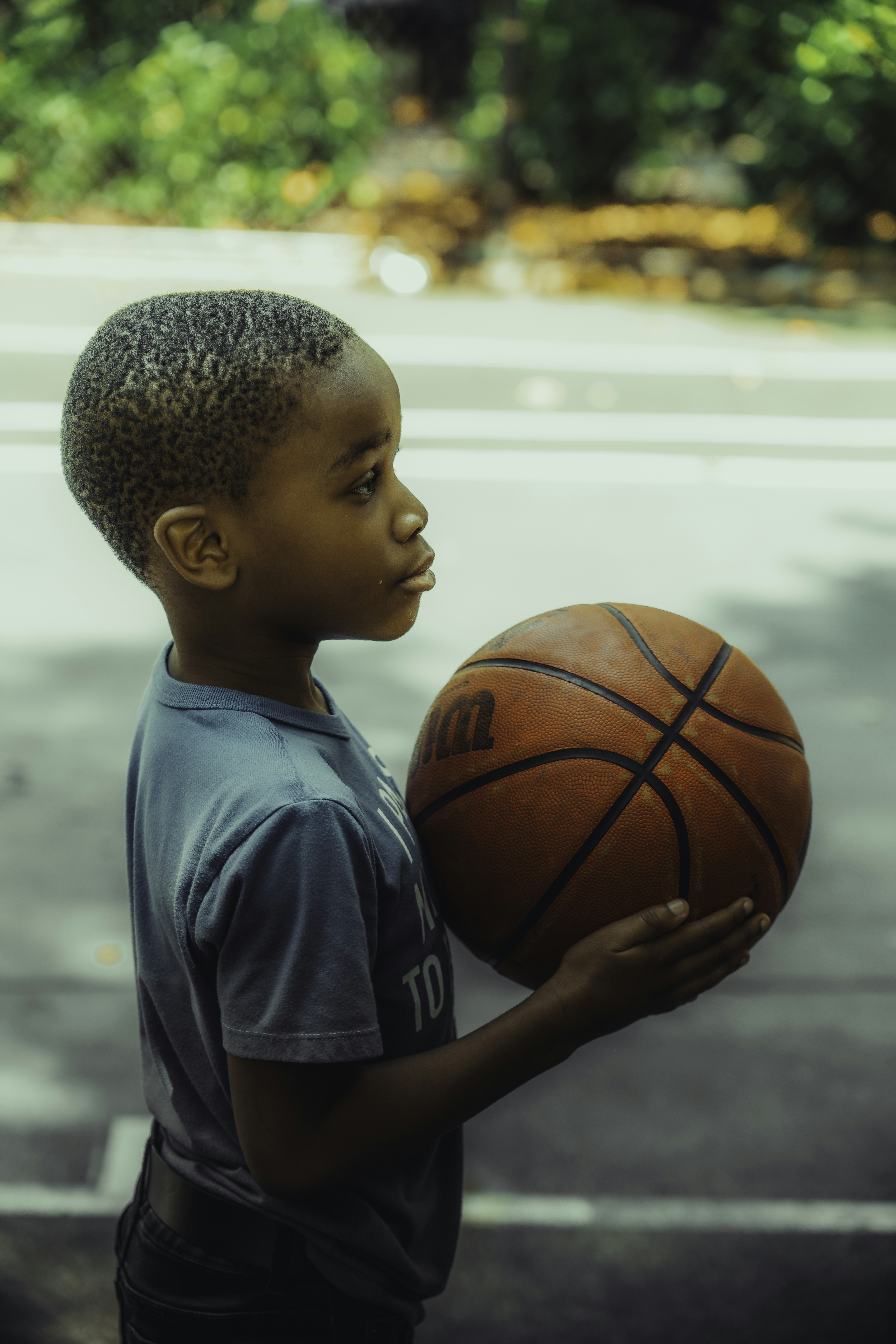 Young boy holding a basketball, gazing thoughtfully while standing on a court. The scene captures a moment of anticipation and focus.