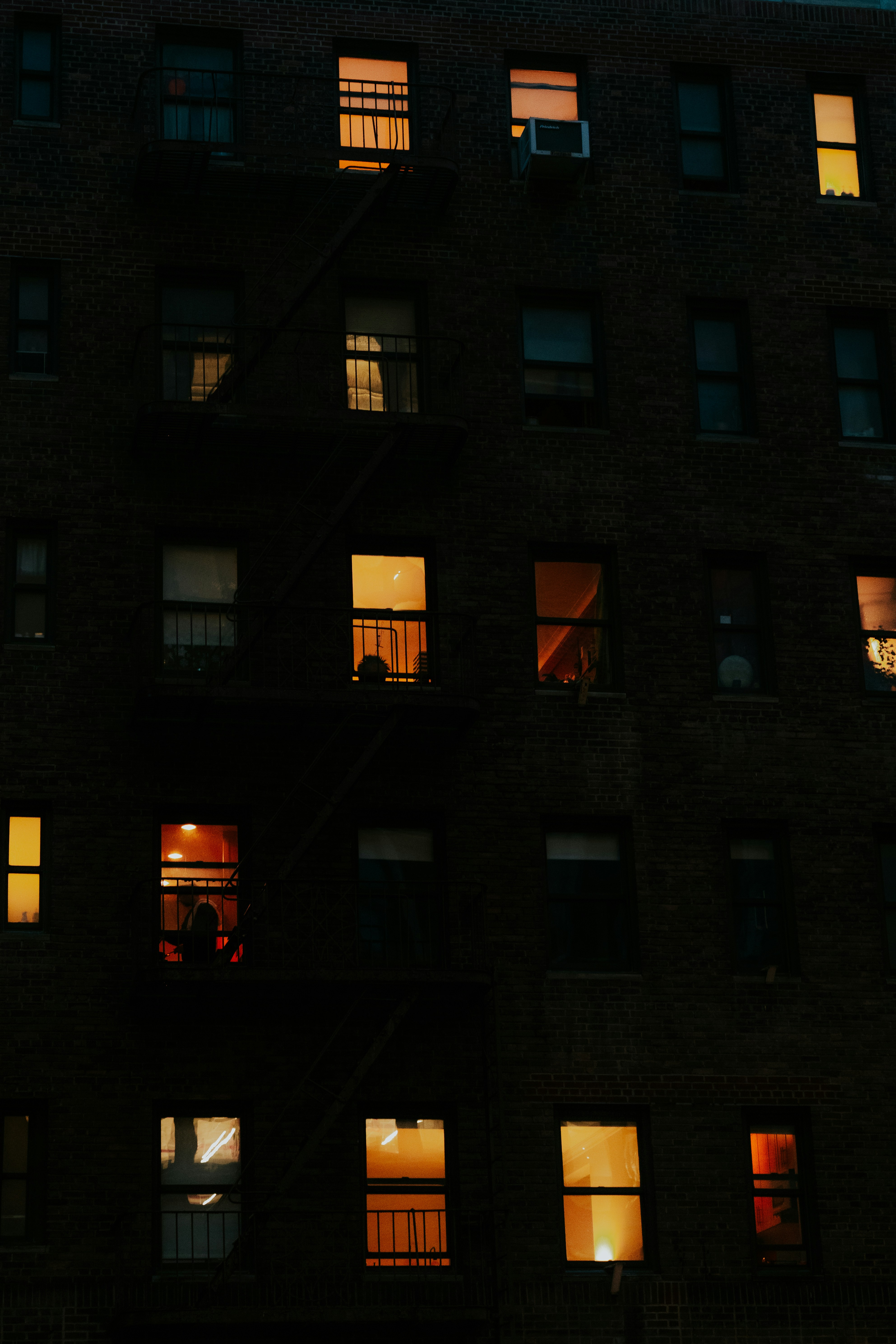 Illuminated windows of a brick apartment building reveal glimpses of life within as night falls.