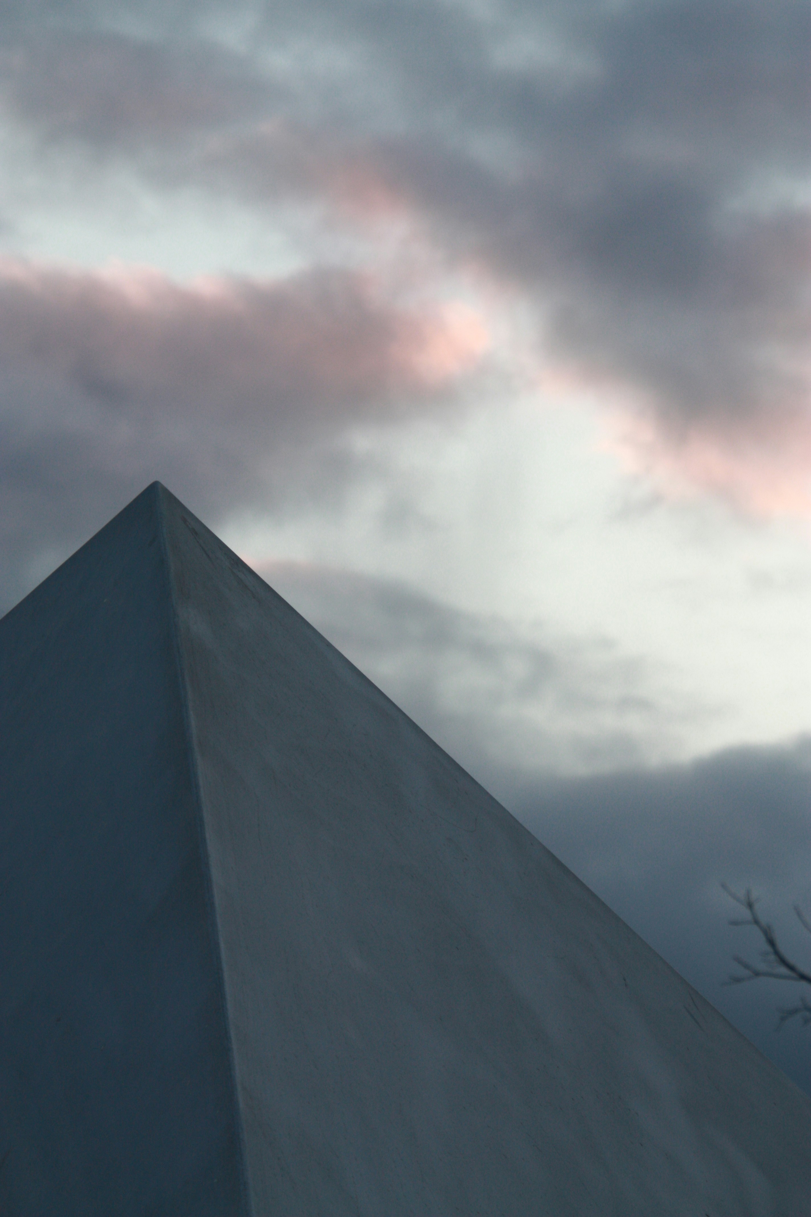 A sharp, angular pyramid structure rises against a backdrop of softly diffused clouds, hinting at the transition from day to night.
