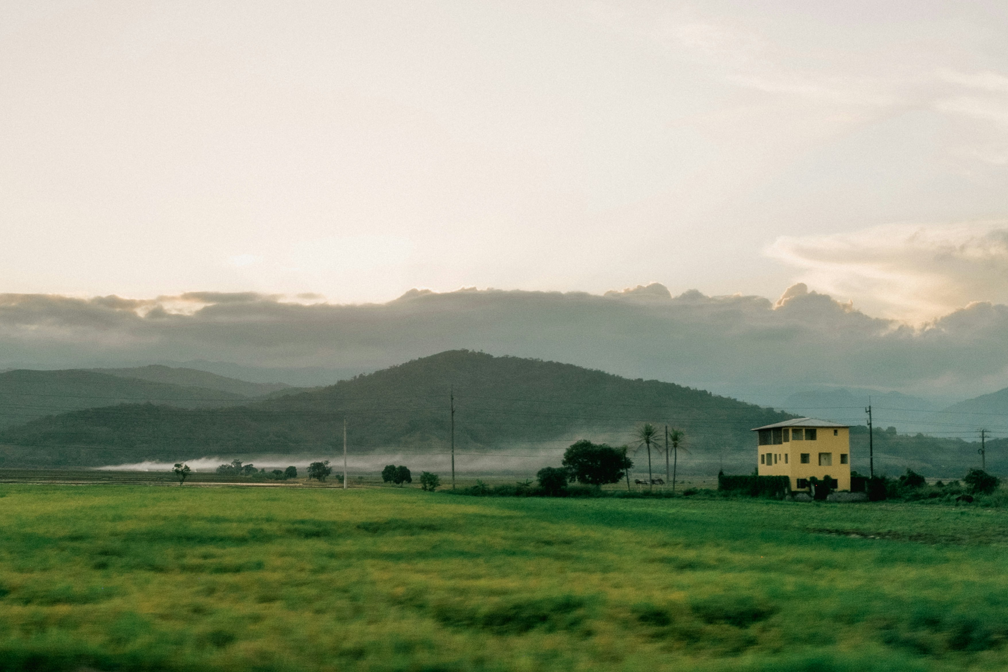 A yellow house stands alone in a lush green field, framed by distant mountains under a soft, pastel sky.