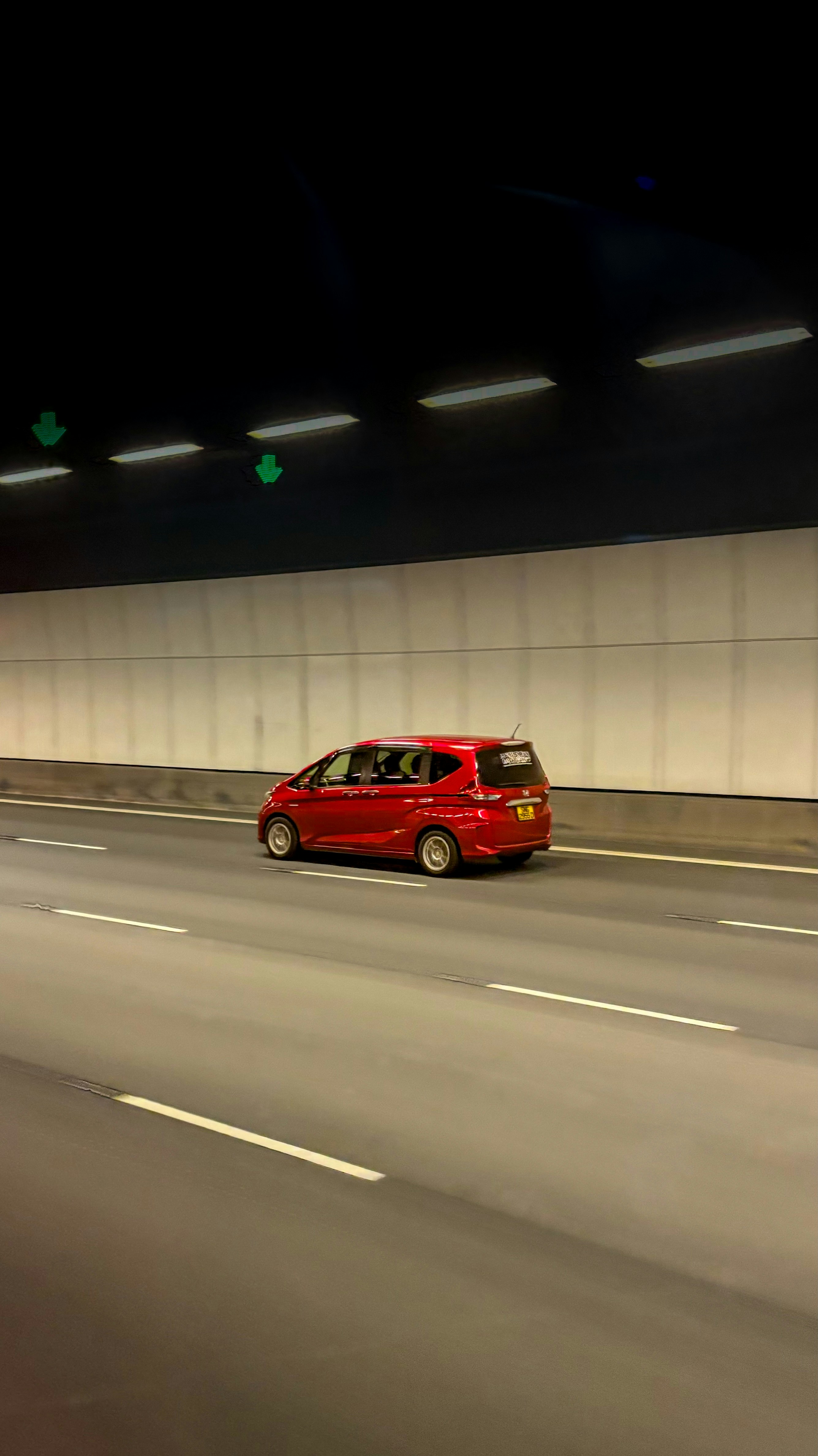 A vibrant red car speeds through a dimly lit tunnel, illuminated by overhead lights and directional signs.