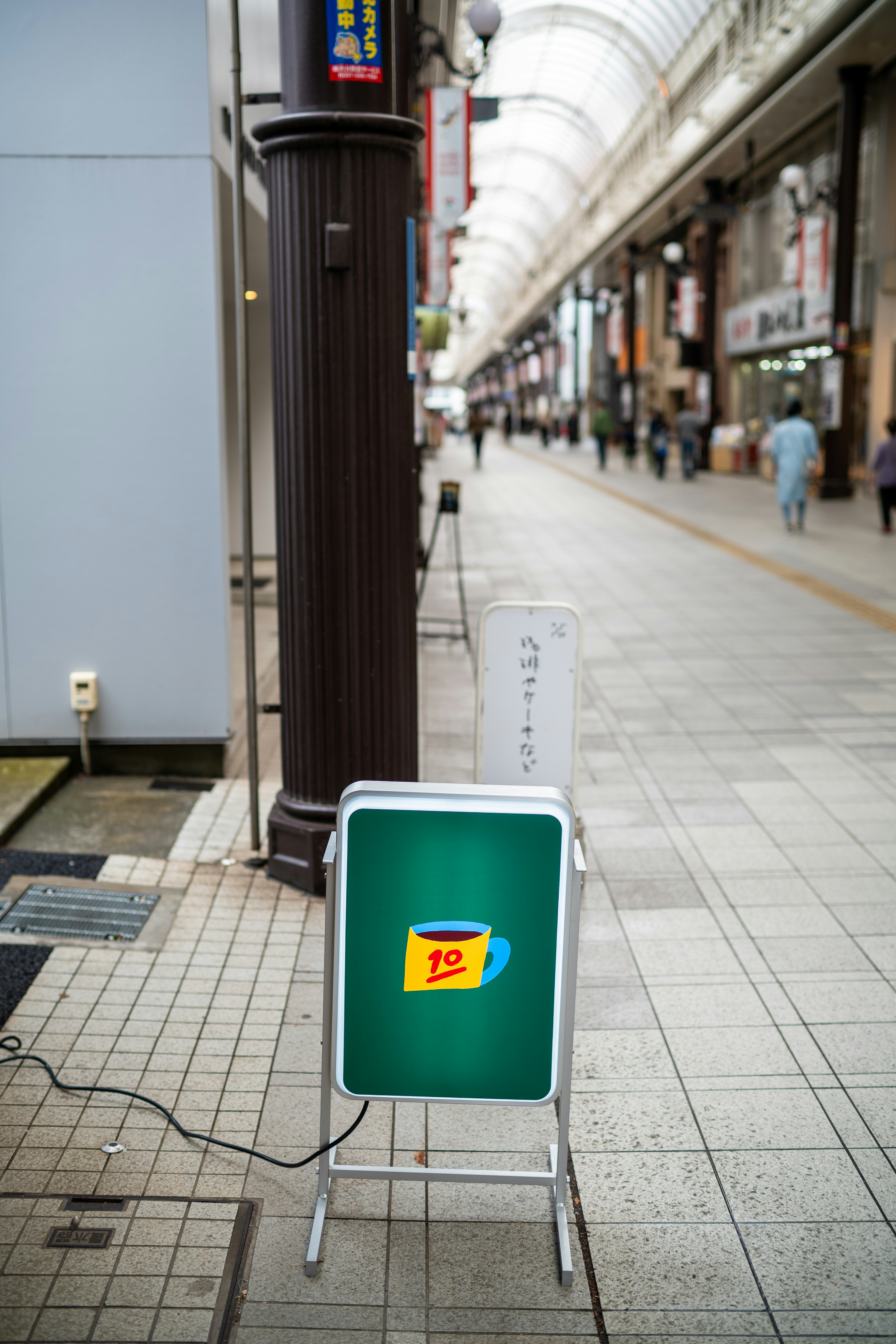 A green sign featuring a coffee cup icon and the number 10 stands on a bustling street, inviting passersby to enjoy a beverage. The setting showcases a lively shopping arcade.