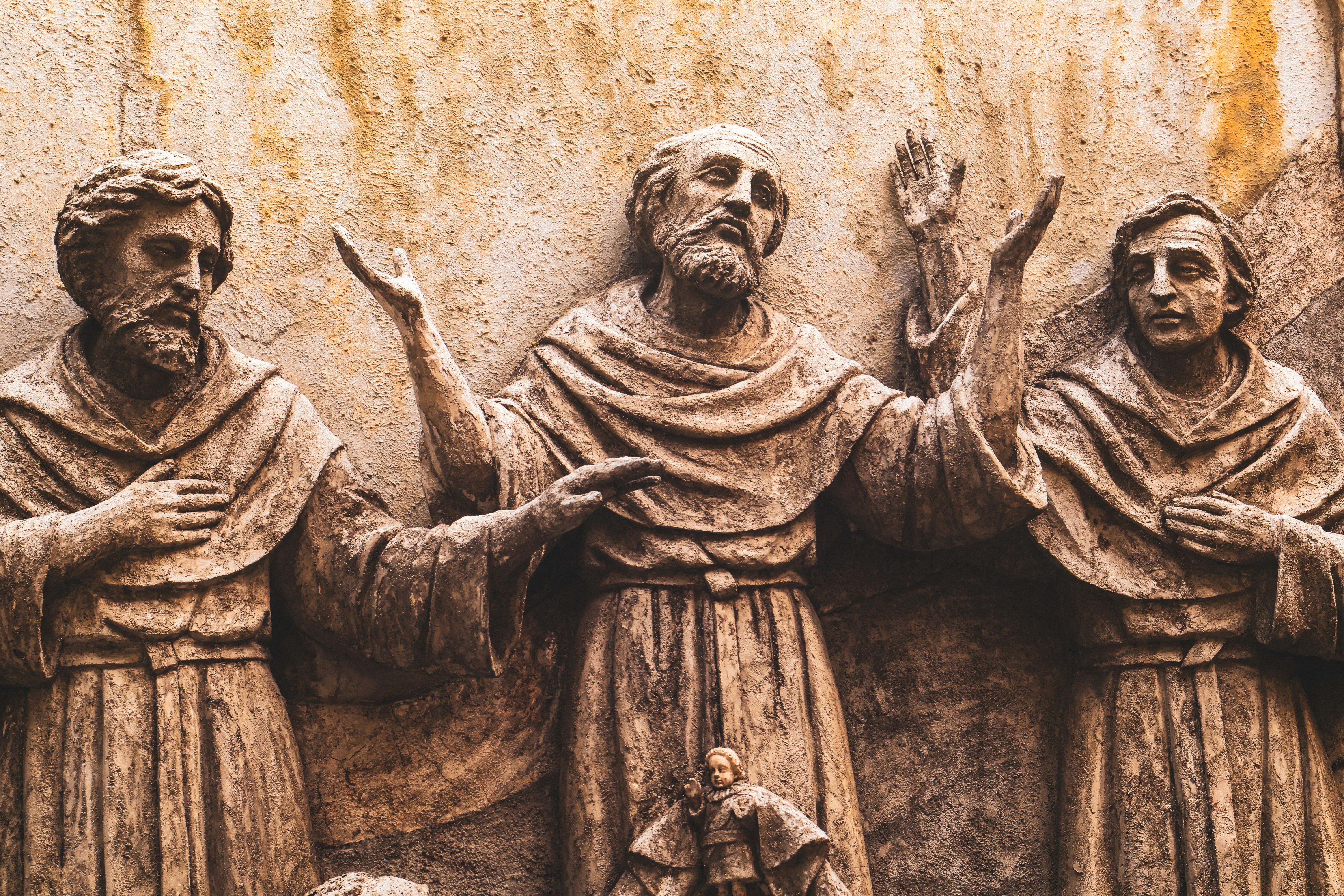 Three intricately carved figures in robes engage in a spiritual conversation, with a small figure at their feet. The texture of the stone adds depth to their expressions.