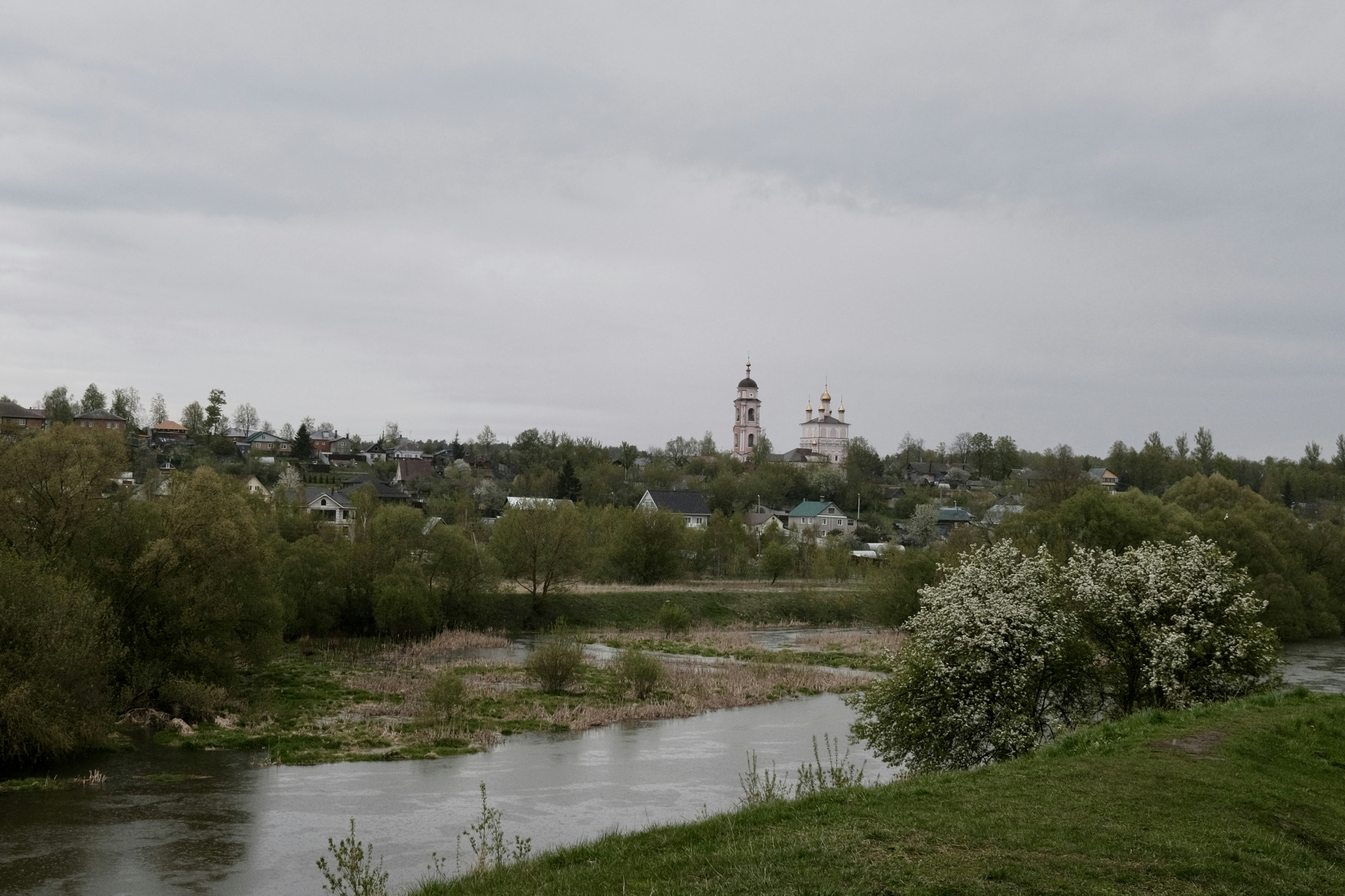 A tranquil riverside scene featuring lush greenery and distant church spires under a cloudy sky. The gentle flow of the water complements the peaceful atmosphere.