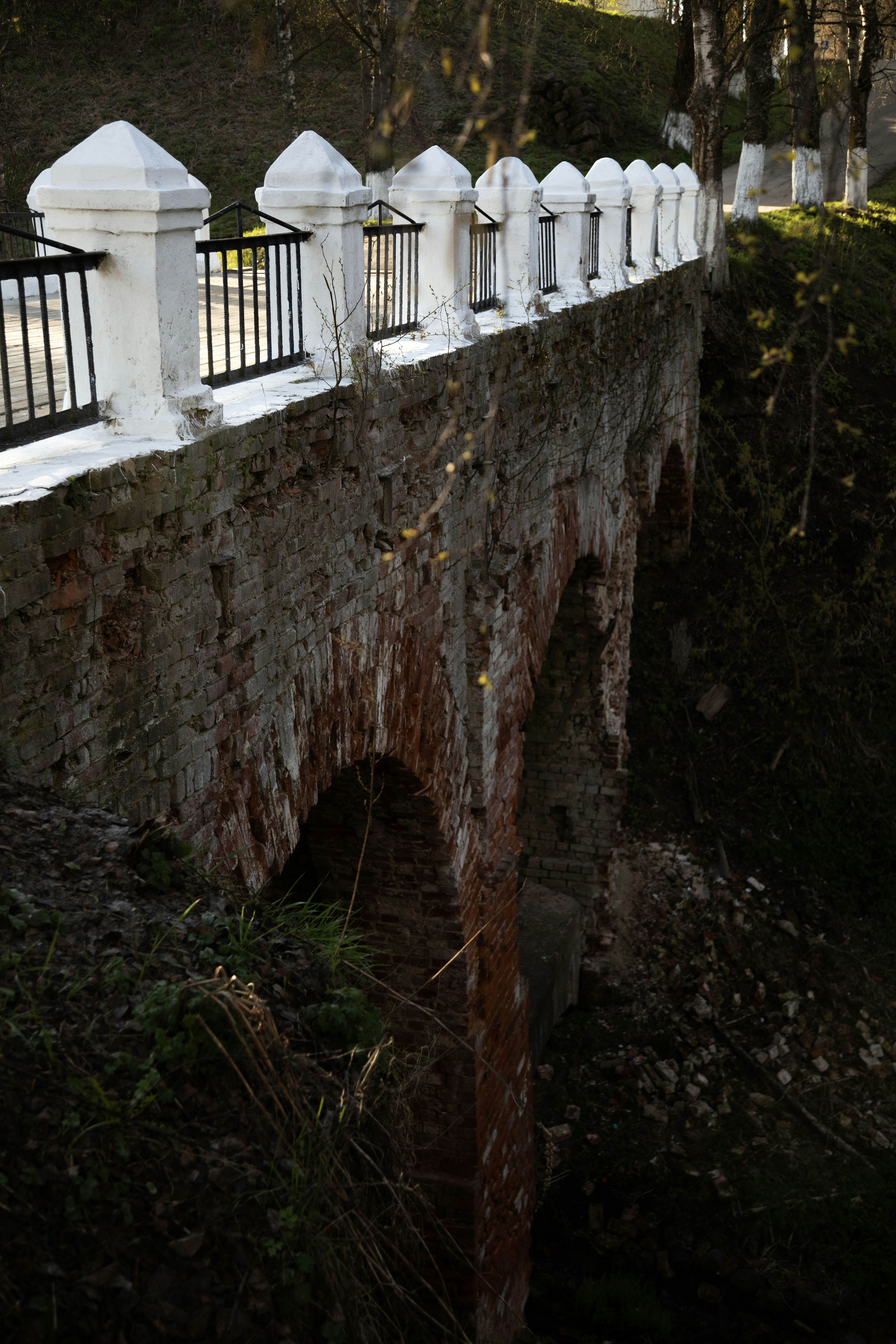 A weathered brick bridge adorned with white balusters spans a ravine, hinting at a rich history. Shadows dance across the structure, enhancing its rustic charm.