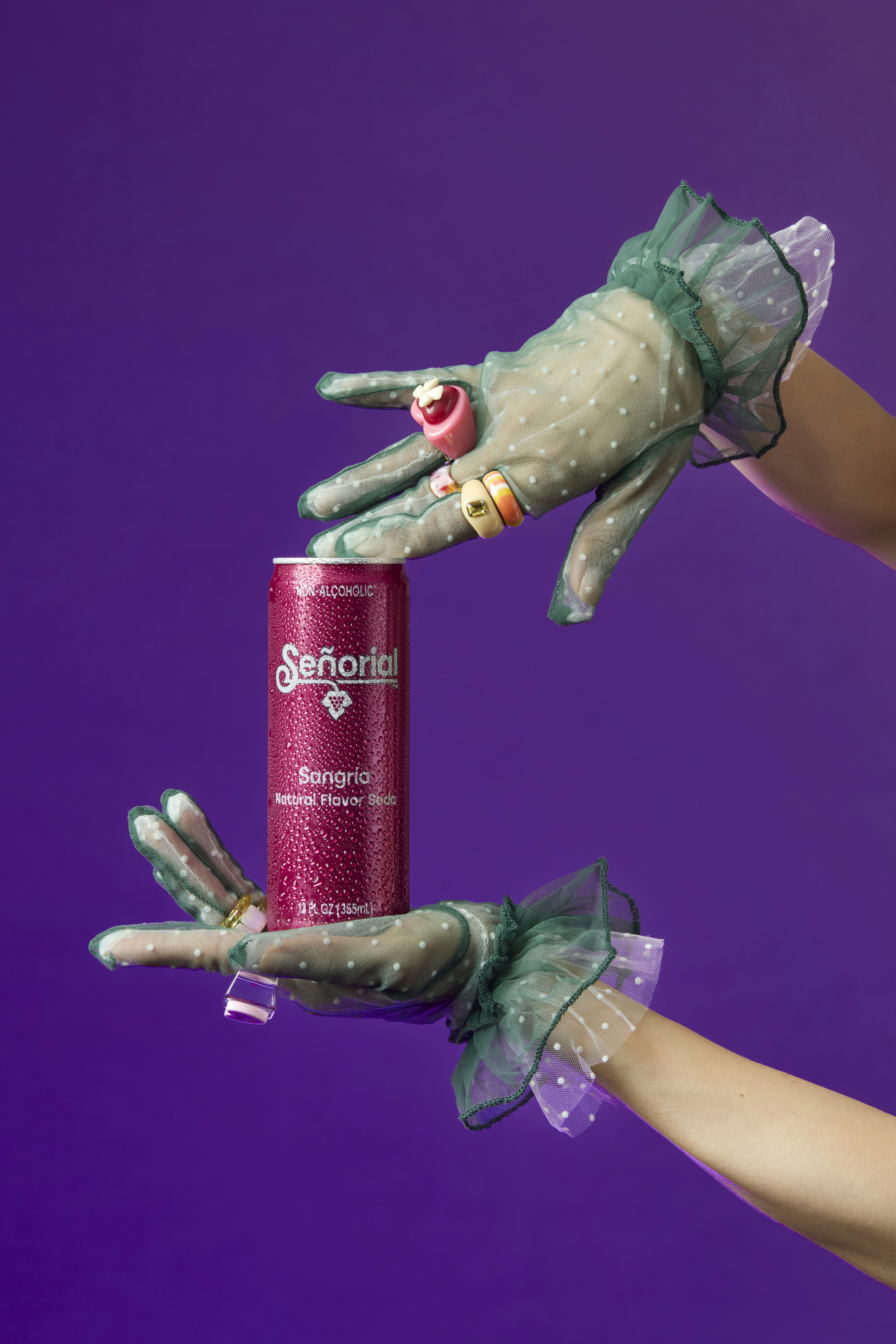 Glimmering Pink Soda Can Balanced Between Sheer Green Gloves on a Purple Studio Backdrop