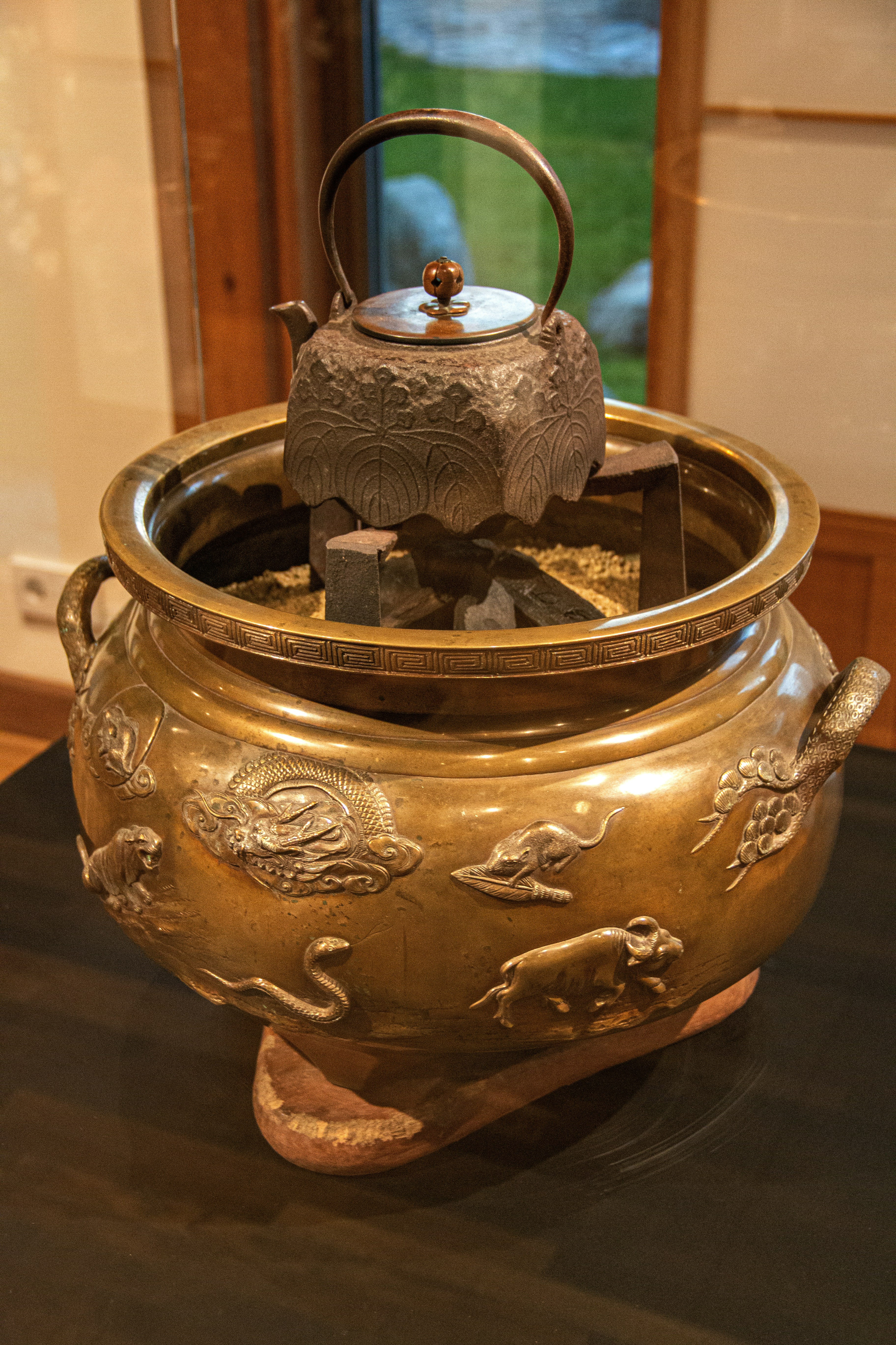 Bronze Embers: An Ornate Teapot Resting on a Sculpted Urn in a Glass Display