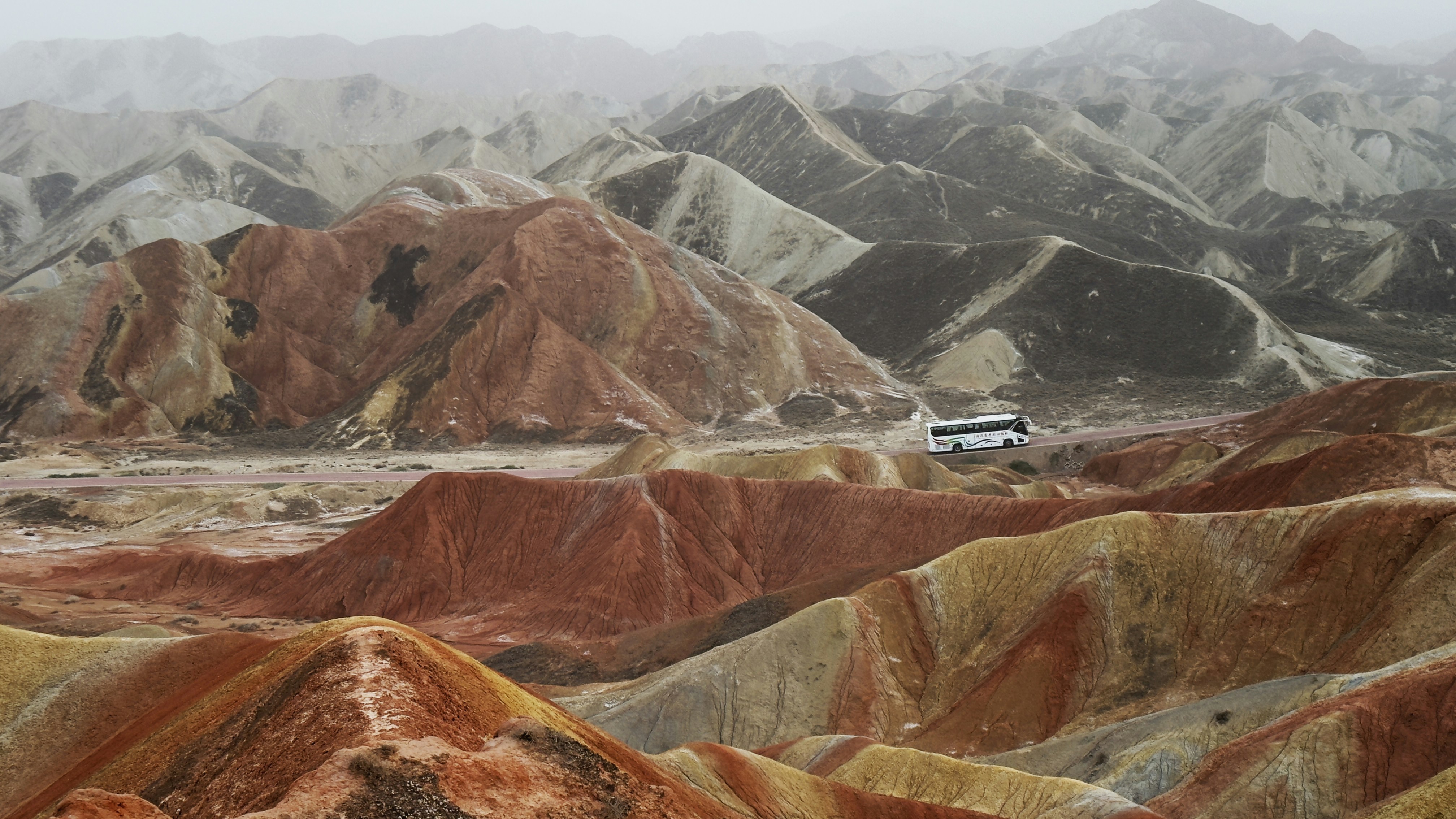 A bus navigates the vibrant, layered hills of a unique geological landscape, showcasing a palette of reds, browns, and yellows. The rugged terrain creates a striking contrast against the soft sky.