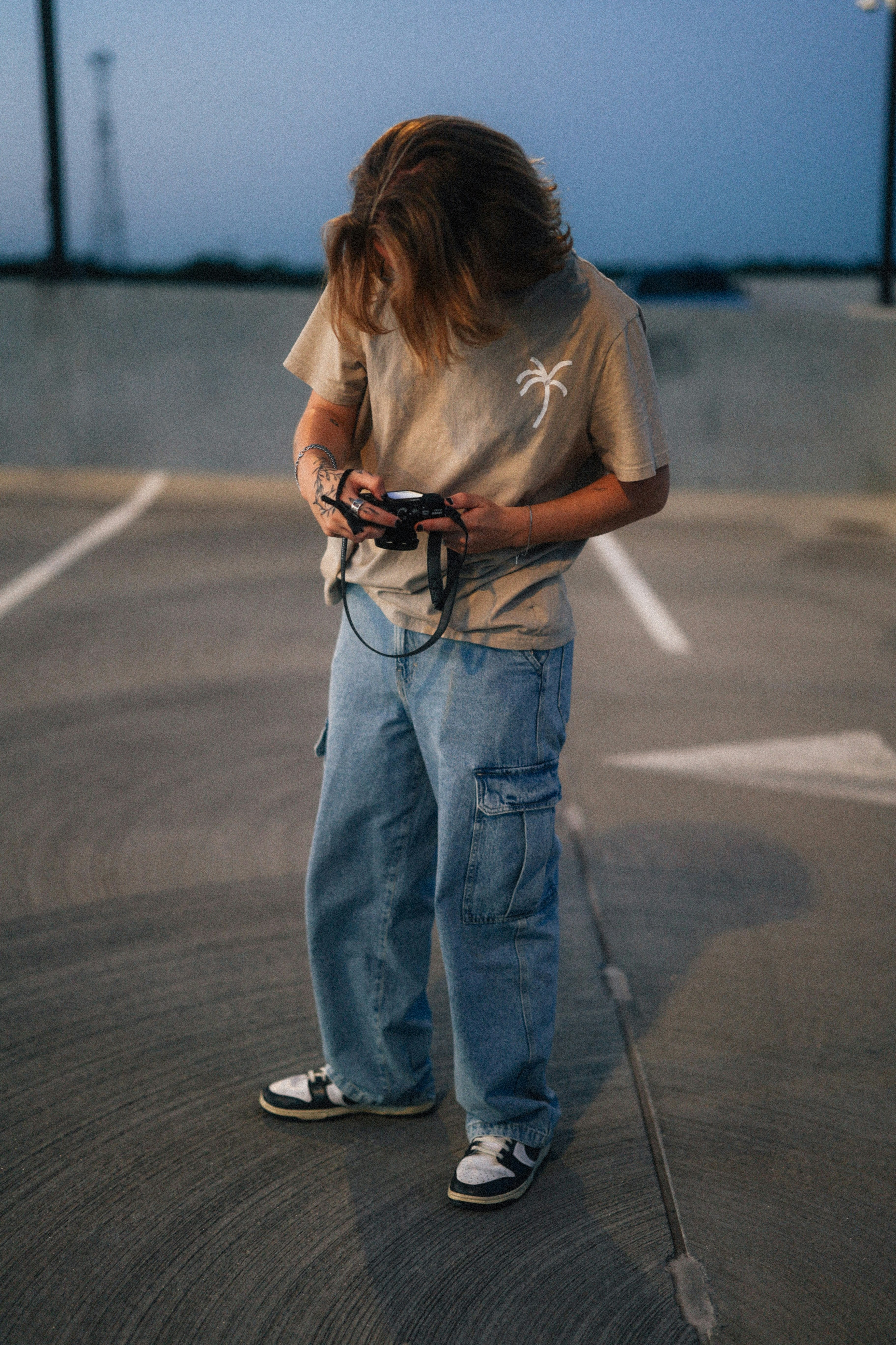 Blue Hour Focus: Palm-Tree Tee Photographer Examines a Handheld Camera in a Quiet Parking Lot