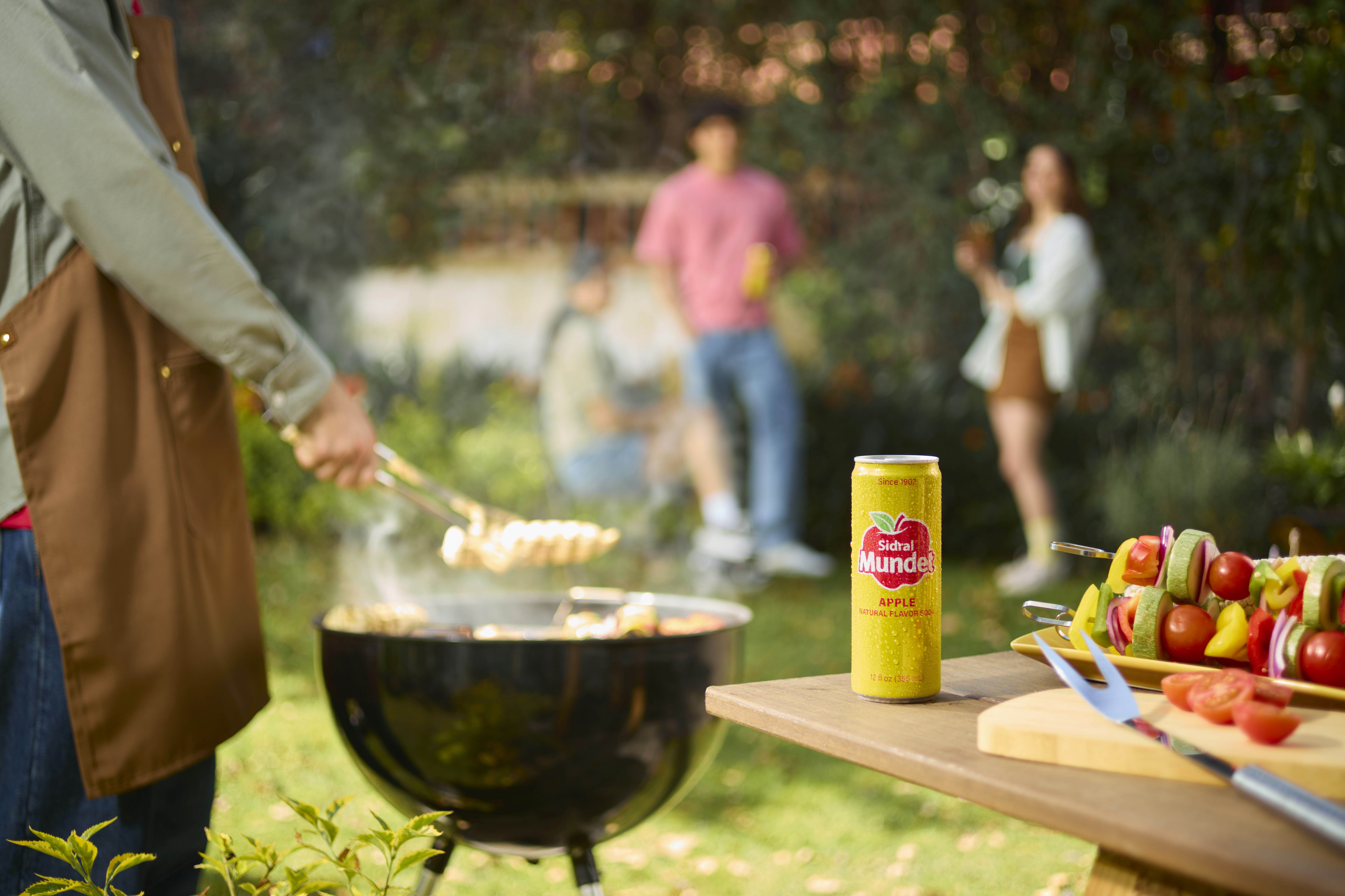 A person grilling skewers at a backyard barbecue, with friends enjoying drinks in the background. A can of beverage is prominently featured on the table.