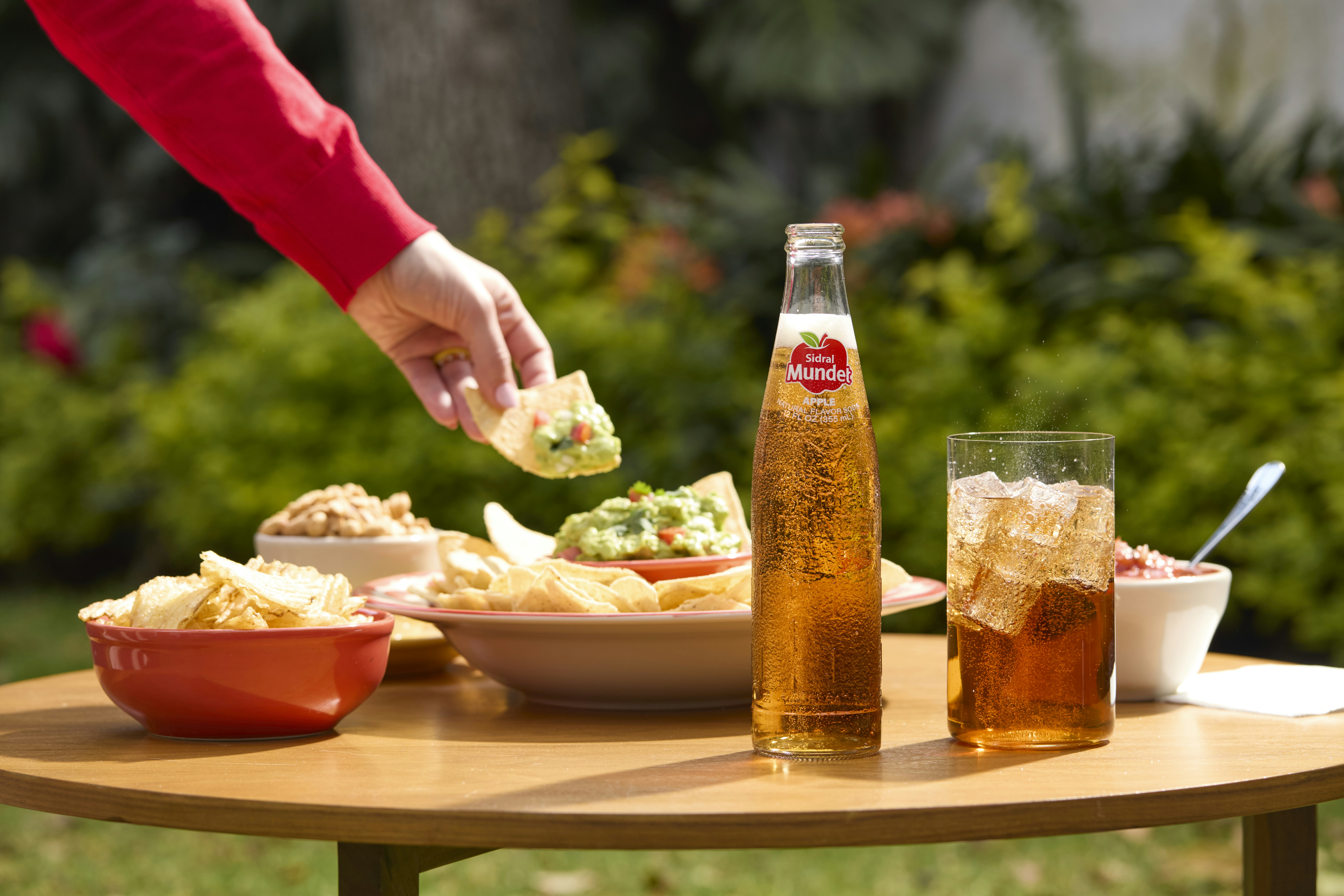 Hand reaching for a chip topped with guacamole, surrounded by various snacks and beverages on a wooden table outdoors.