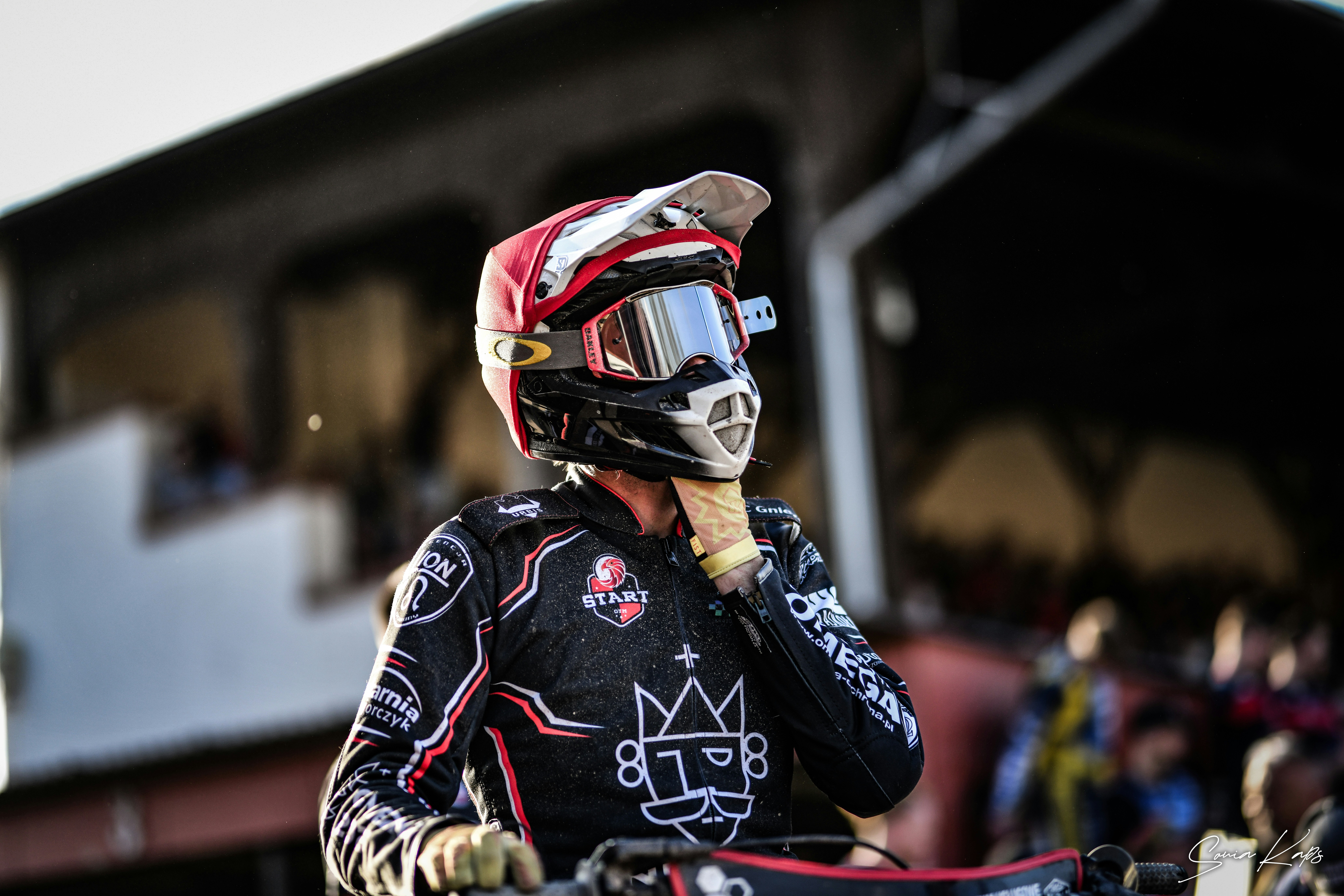Motocross rider in gear, pausing with a thoughtful expression before the race. The background shows a lively crowd at a motocross event.