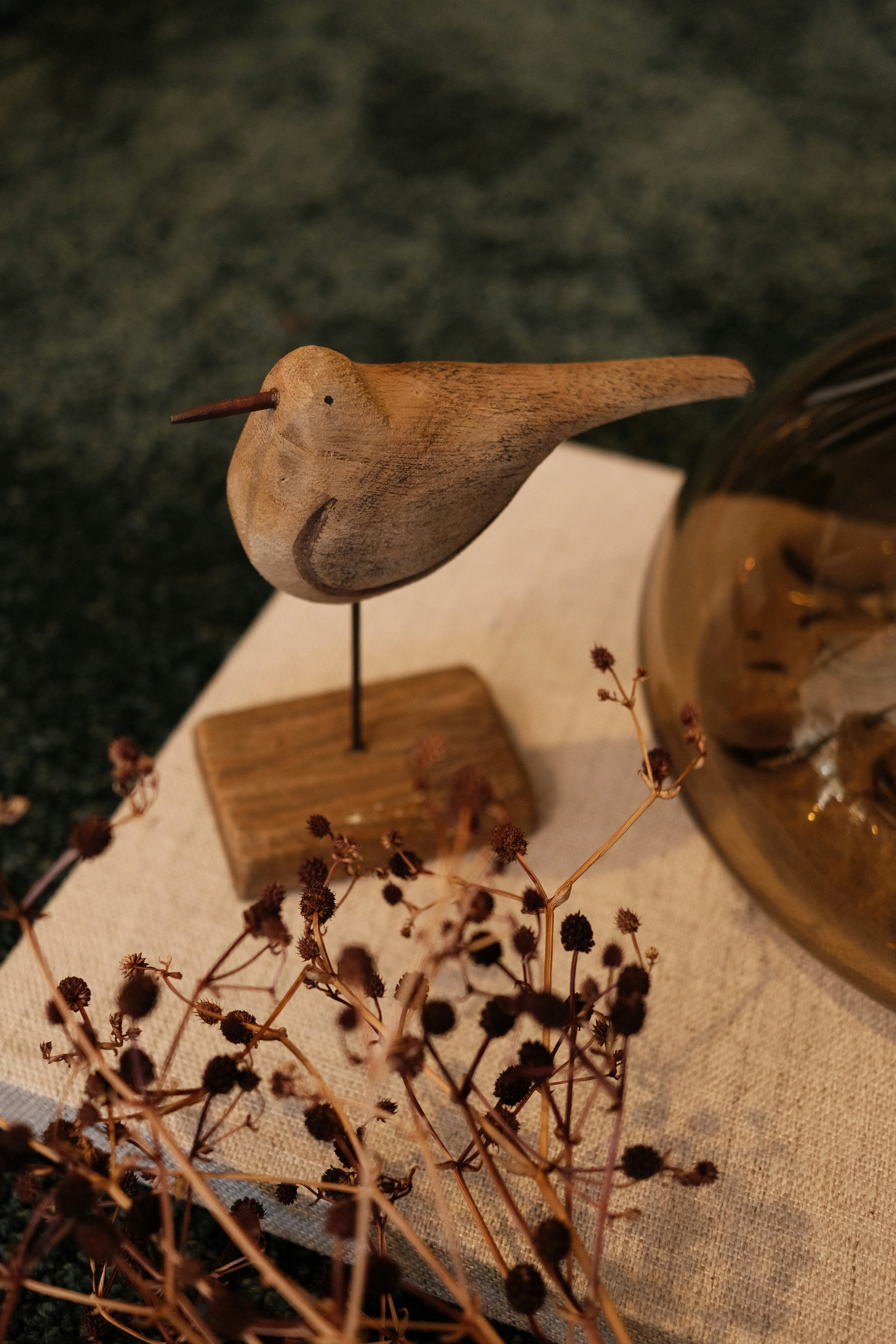Lilt of Wood: A Whittled Bird on a Stand Among Dried Blooms and a Glass Vessel