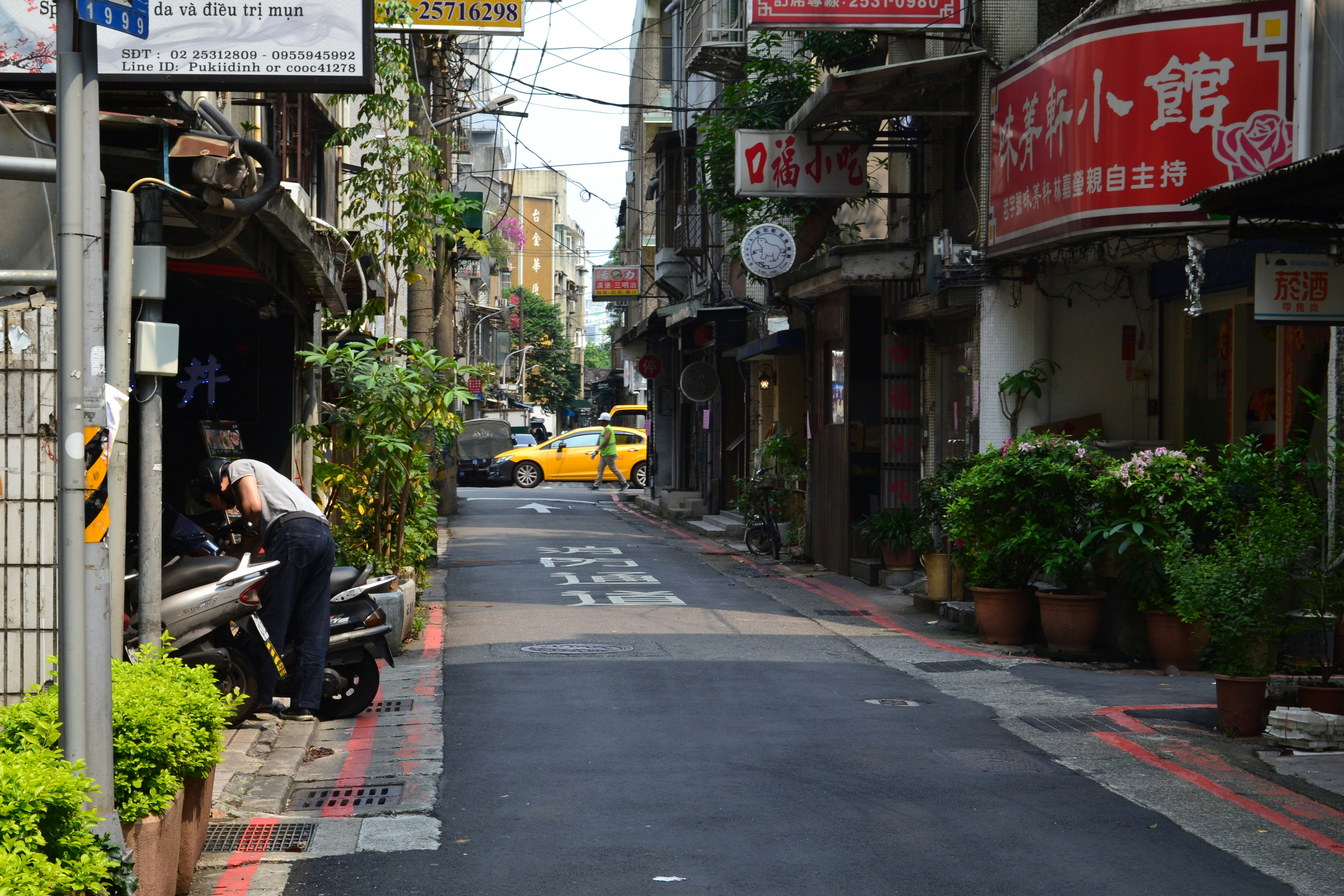 A narrow alleyway in a bustling city, featuring a person working on a scooter surrounded by vibrant greenery and signage. A yellow taxi is visible in the background.