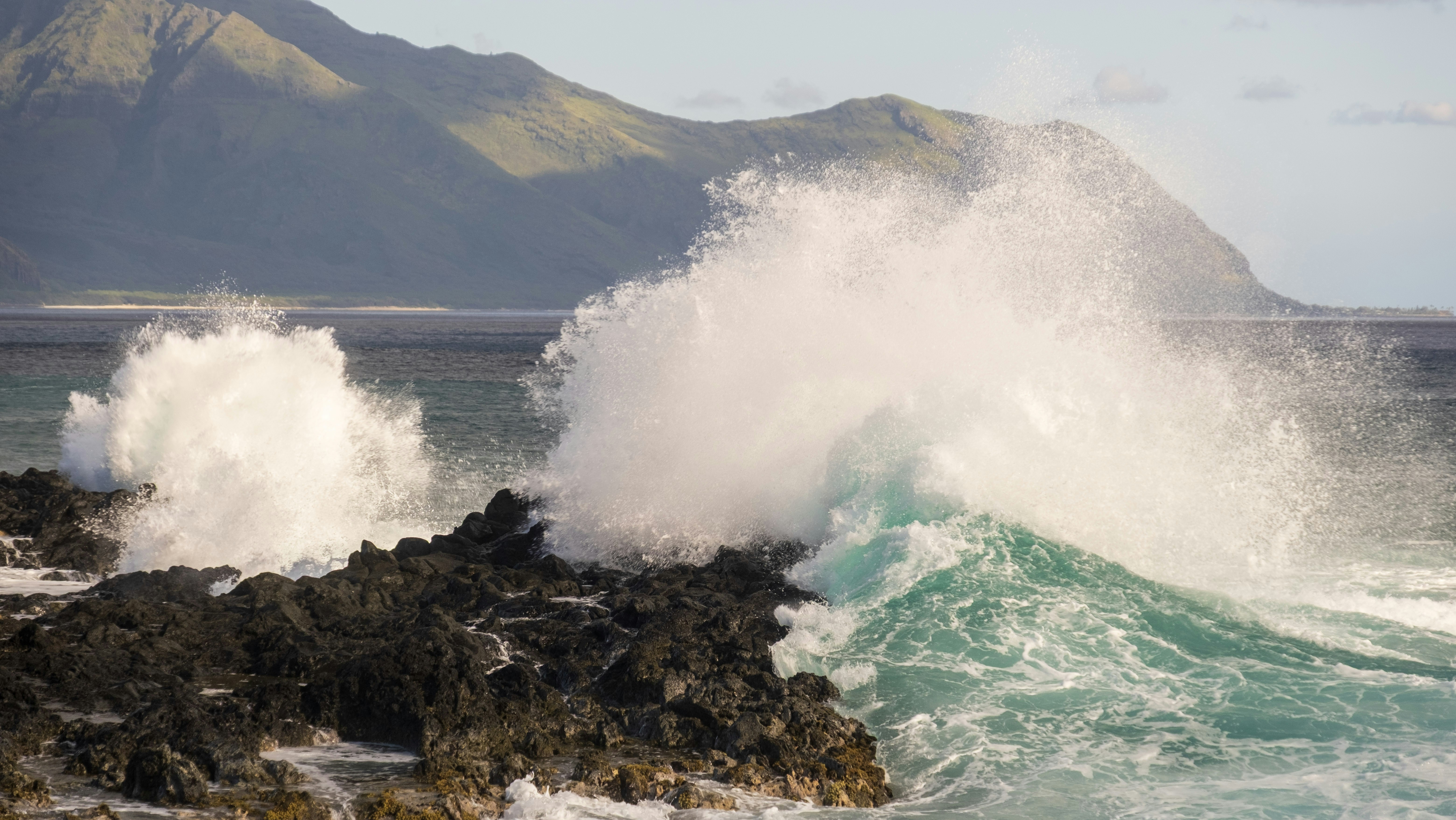 Powerful waves crashing against rugged rocks with a backdrop of lush mountains under a clear sky.