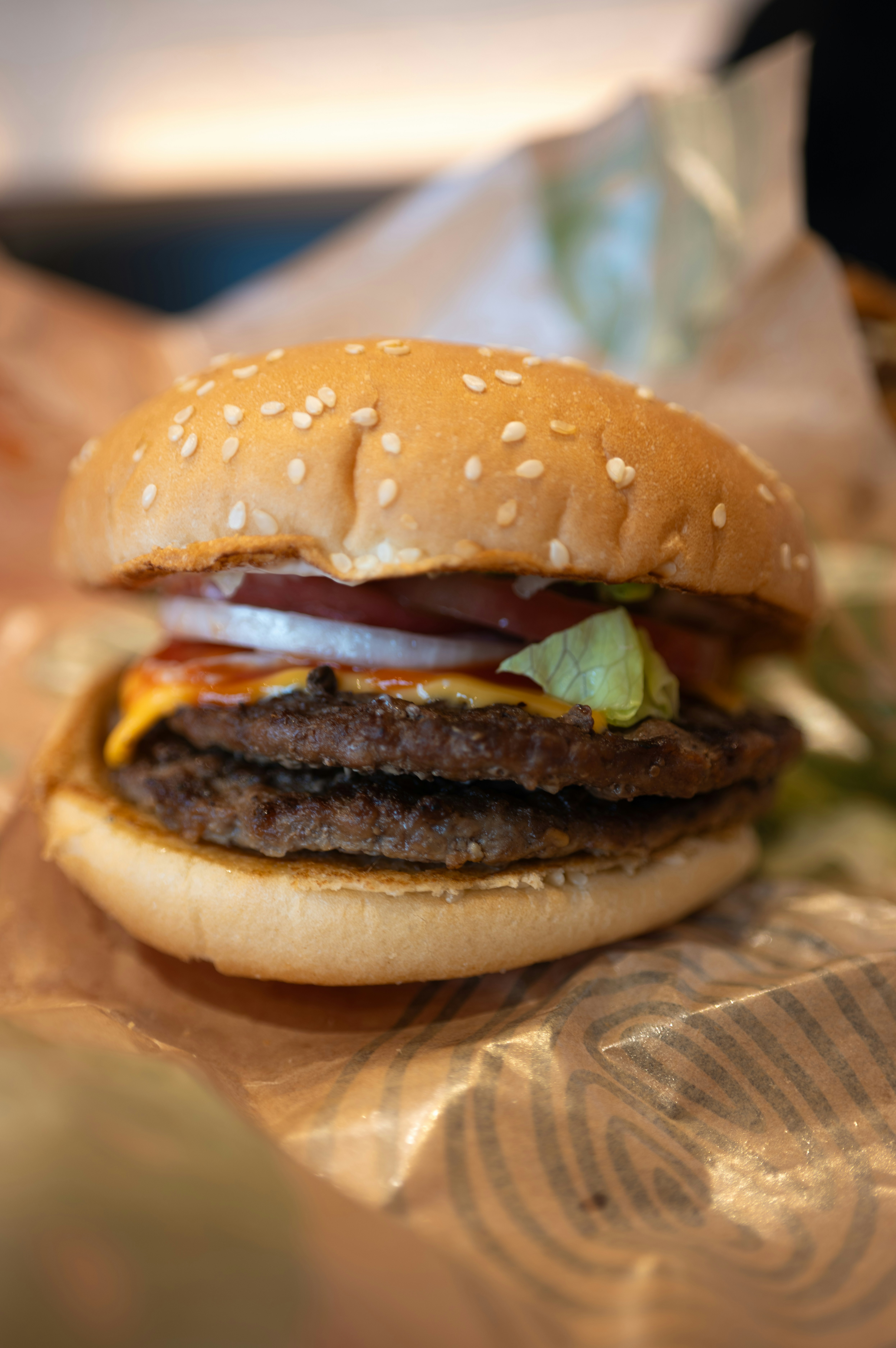 A close-up view of a sesame seed bun burger featuring two juicy patties, melted cheese, fresh lettuce, and slices of tomato, set against a blurred background.
